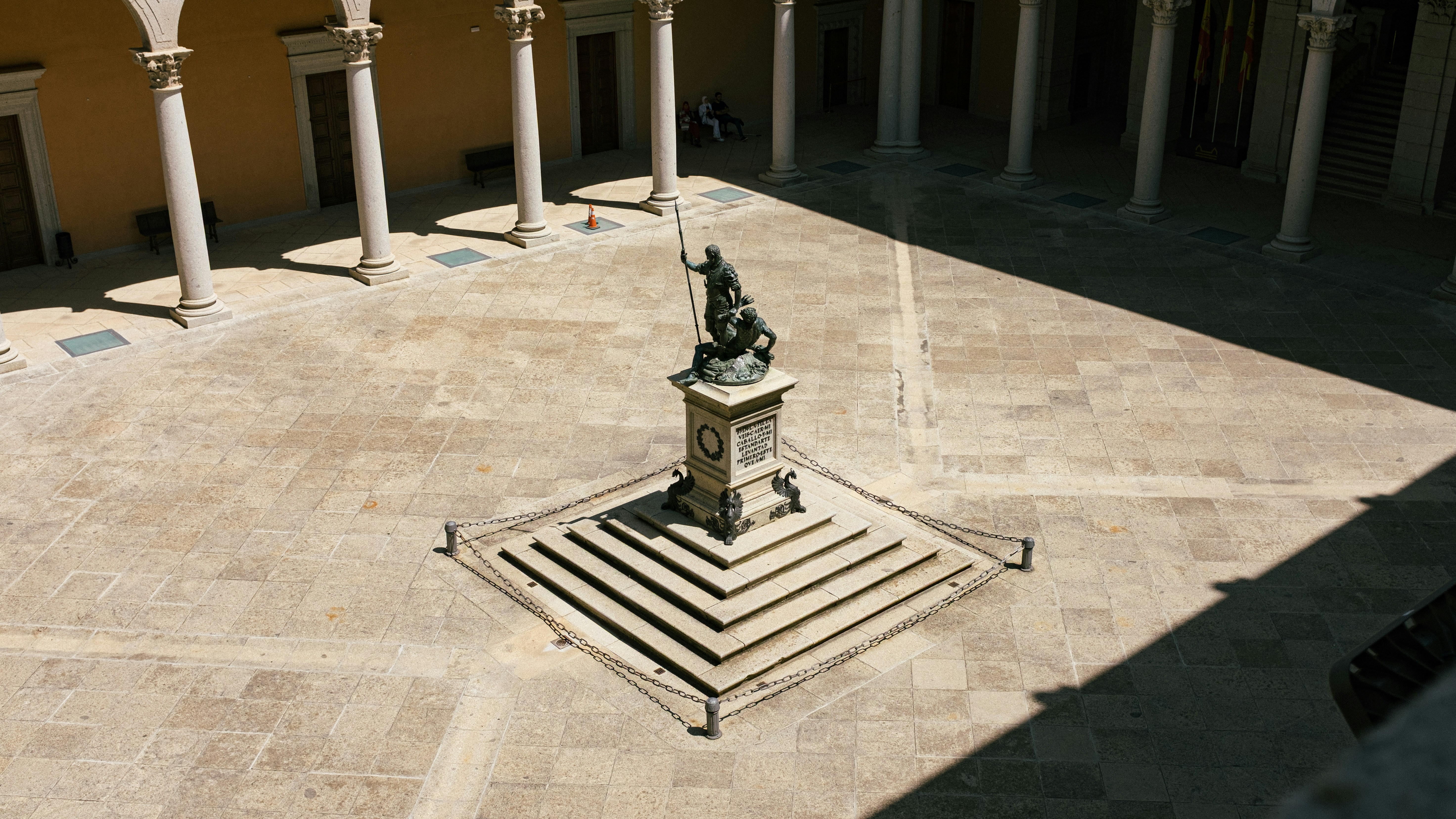 A statue of a warrior stands proudly on a stepped pedestal in a sunlit courtyard, surrounded by columns and shadowy corners.