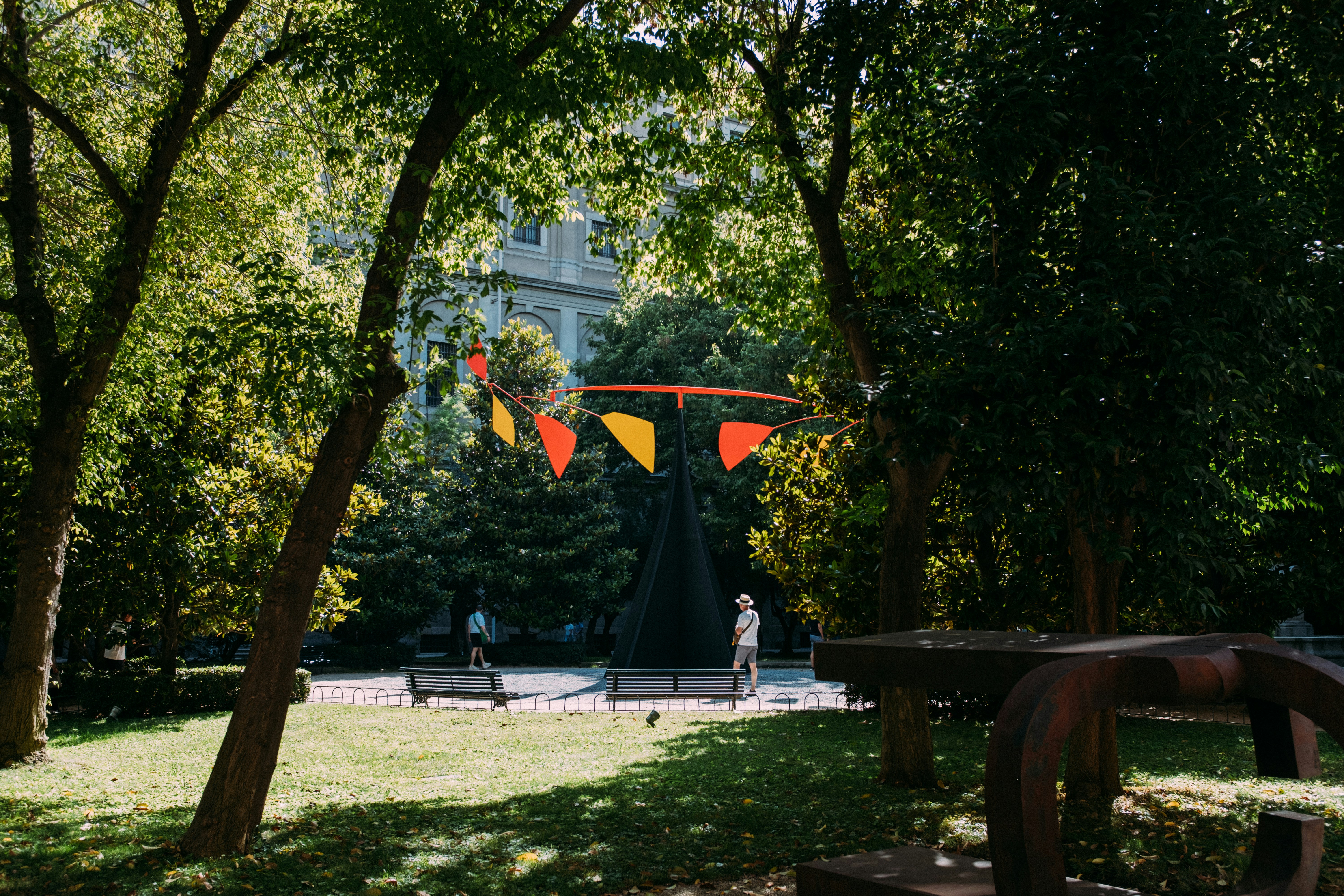 Colorful flags sway gently above a modern sculpture, framed by lush greenery in a serene park setting.
