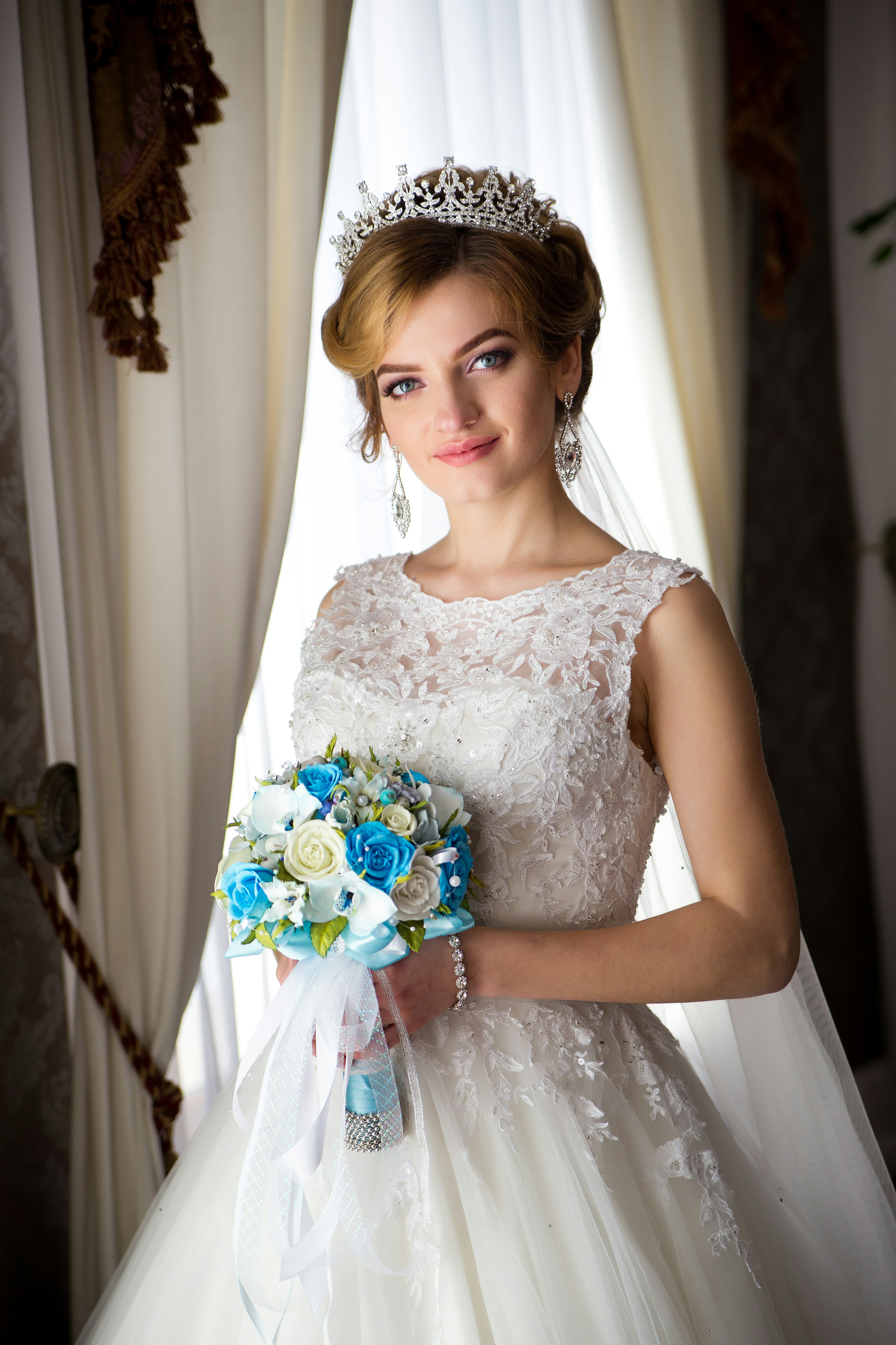 Bride in a beautifully detailed wedding dress holding a bouquet of blue and white flowers, set against an elegant backdrop of draped curtains.