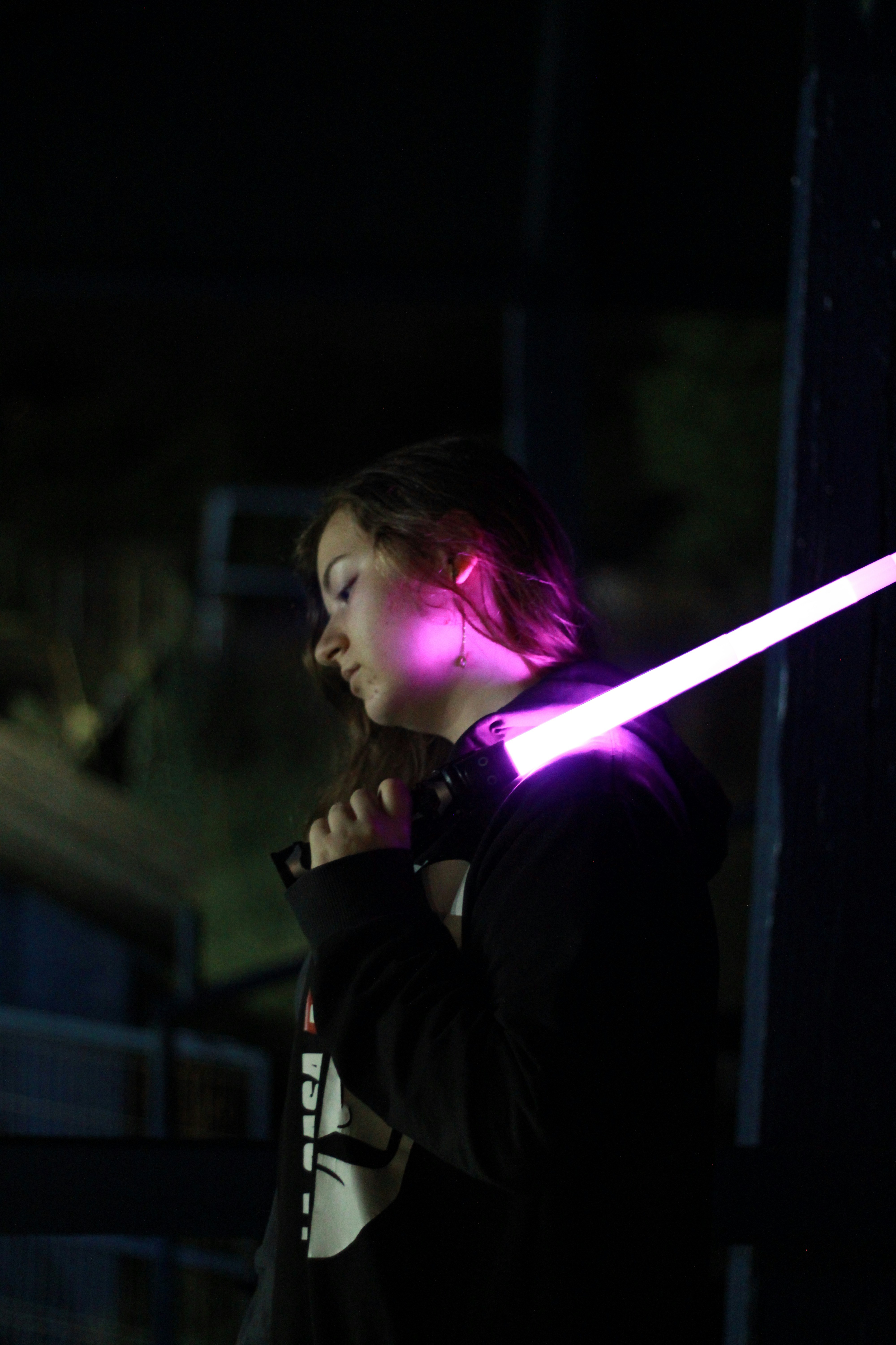 Young woman with a glowing purple lightsaber, lost in thought against a dark backdrop. The contrast highlights her serene expression.