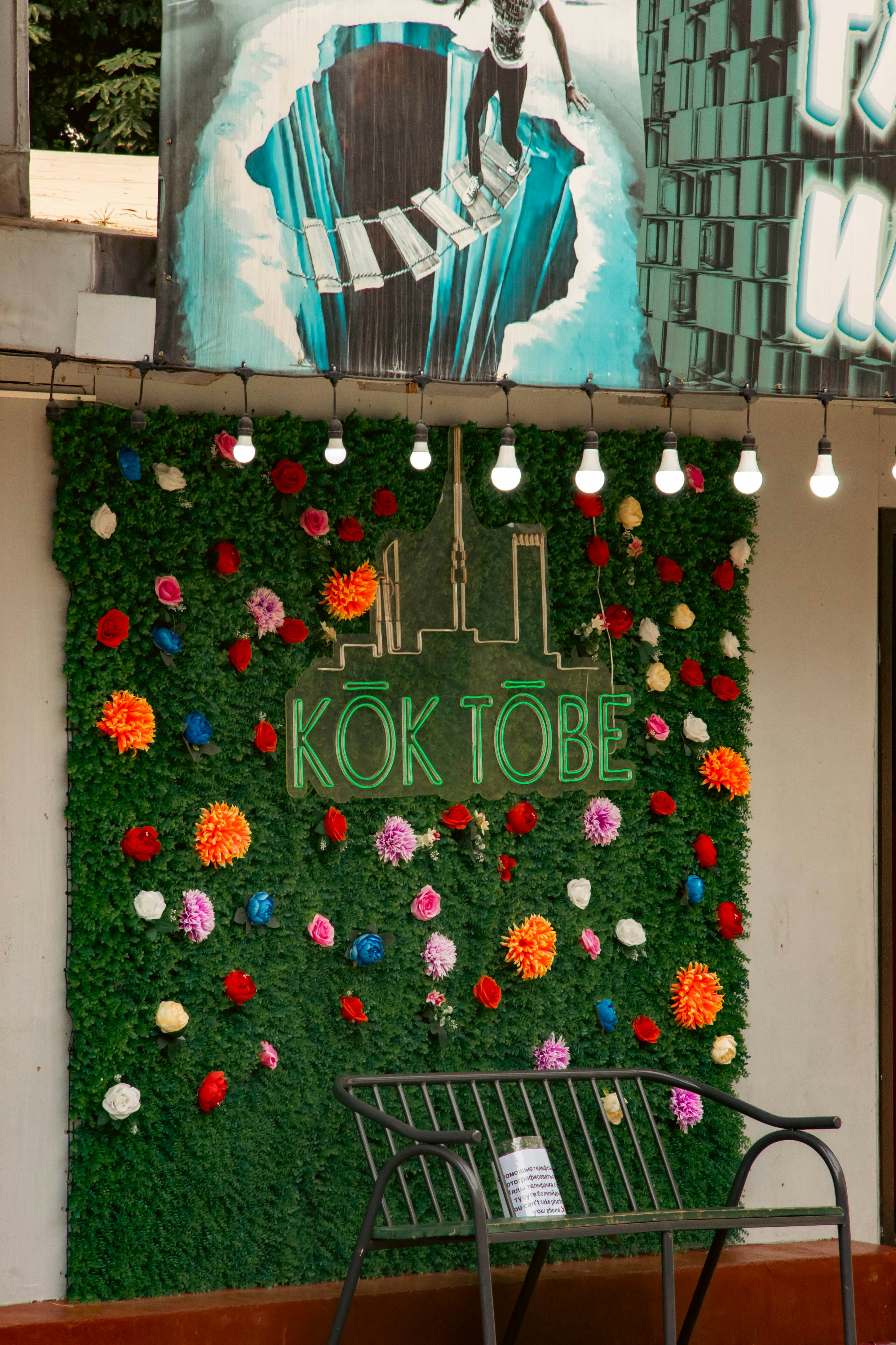 Lush green wall adorned with colorful flowers and the Kök Tobe logo, creating a lively atmosphere. Decorative lights enhance the visual appeal.