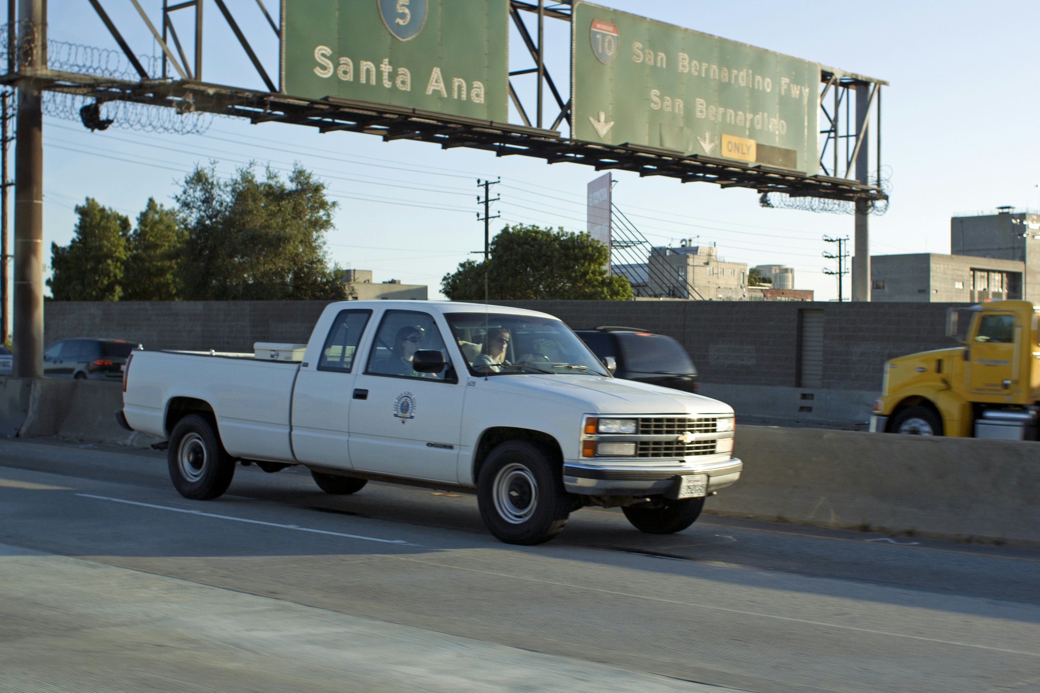 A white pickup truck drives under freeway signs.