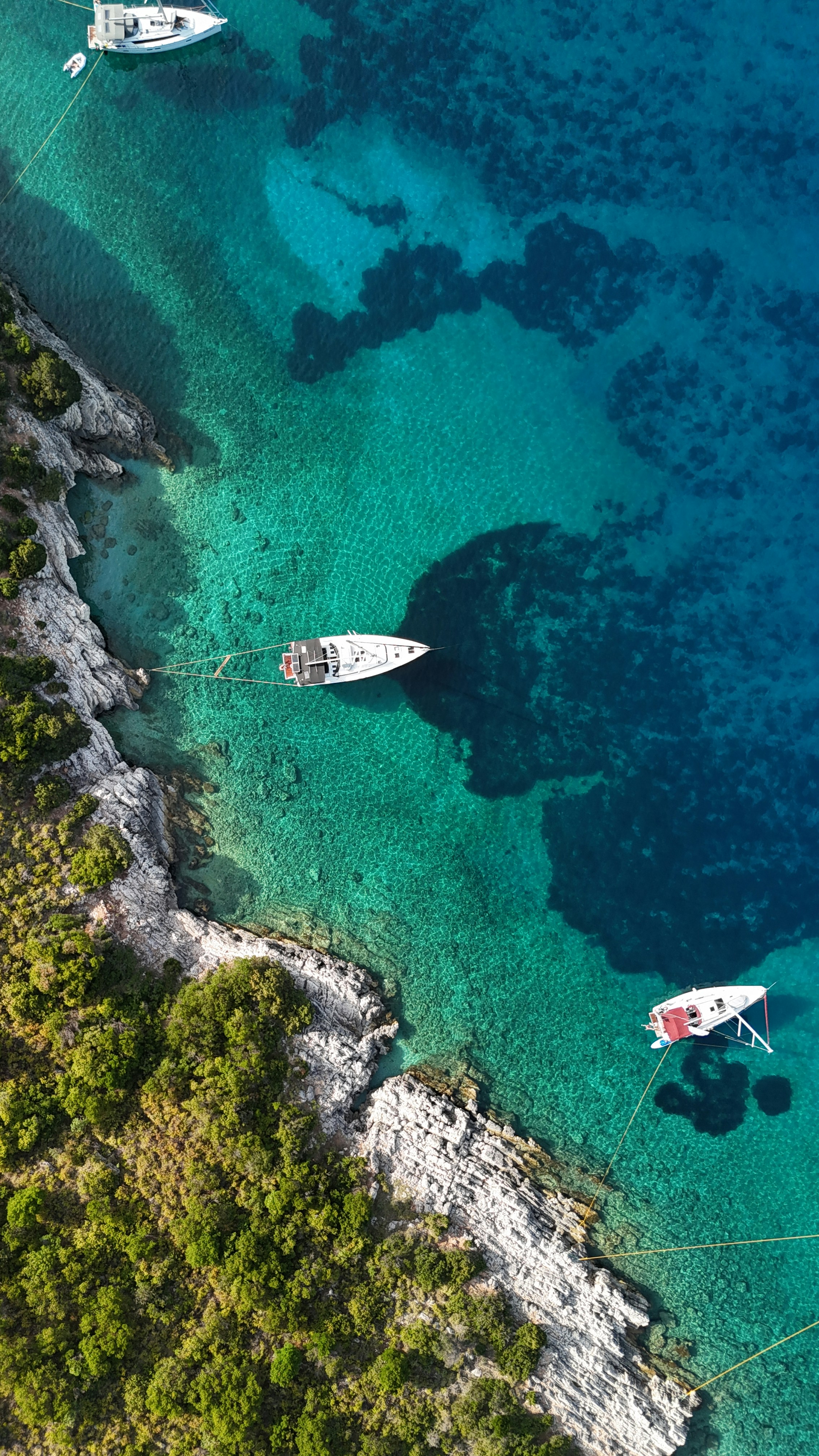 Three boats anchored near a rocky shore with vibrant turquoise waters and lush greenery. Aerial view highlights the serene coastal landscape.