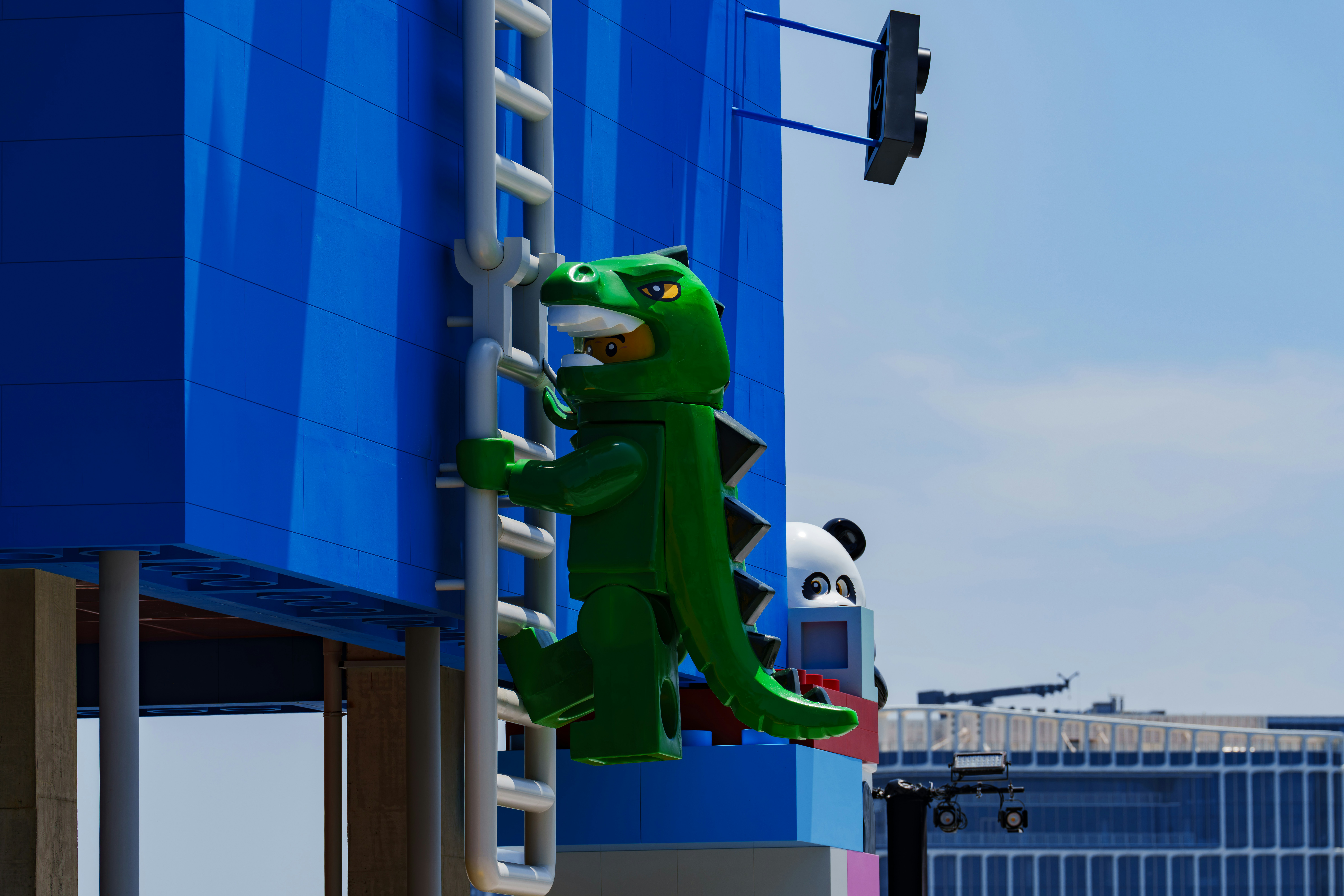 A green LEGO dinosaur character climbs a ladder on a colorful building, with a panda figure peeking from behind. The playful scene captures the essence of imaginative play.
