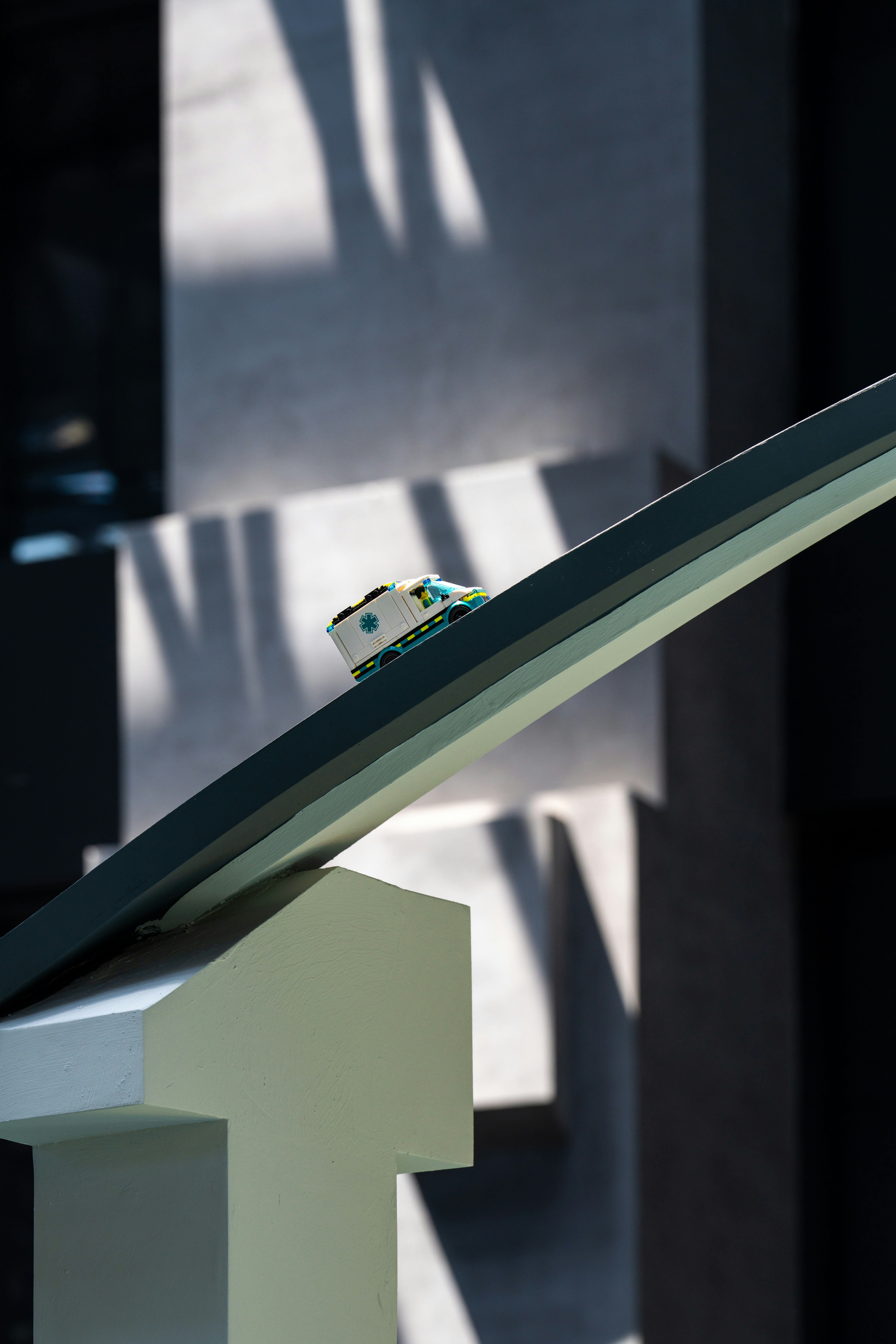 A toy ambulance navigates a curved ramp, framed by dramatic shadows and geometric shapes in the background.