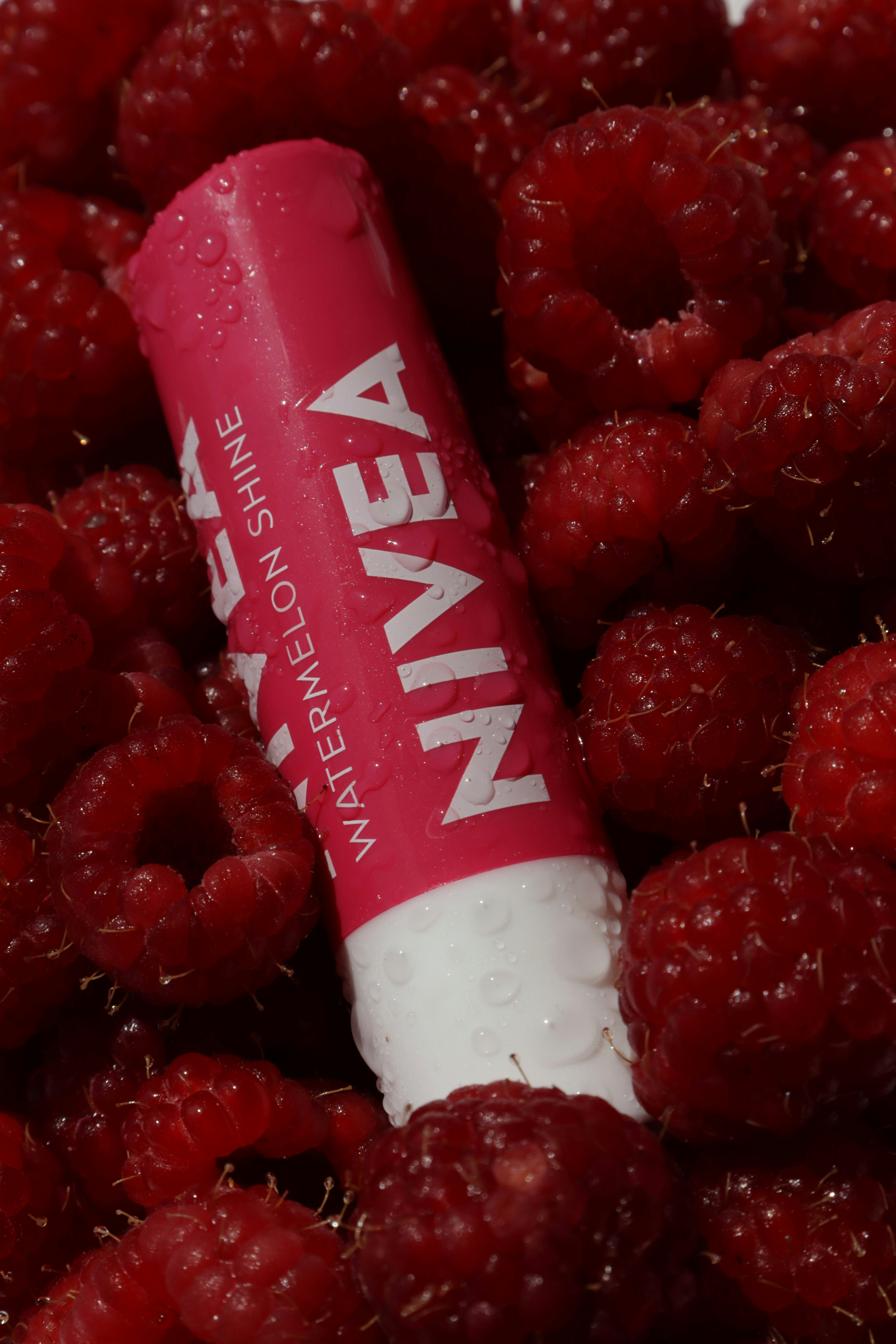NIVEA Watermelon Shine lip balm nestled among fresh, juicy raspberries, showcasing a vibrant color contrast and inviting texture.