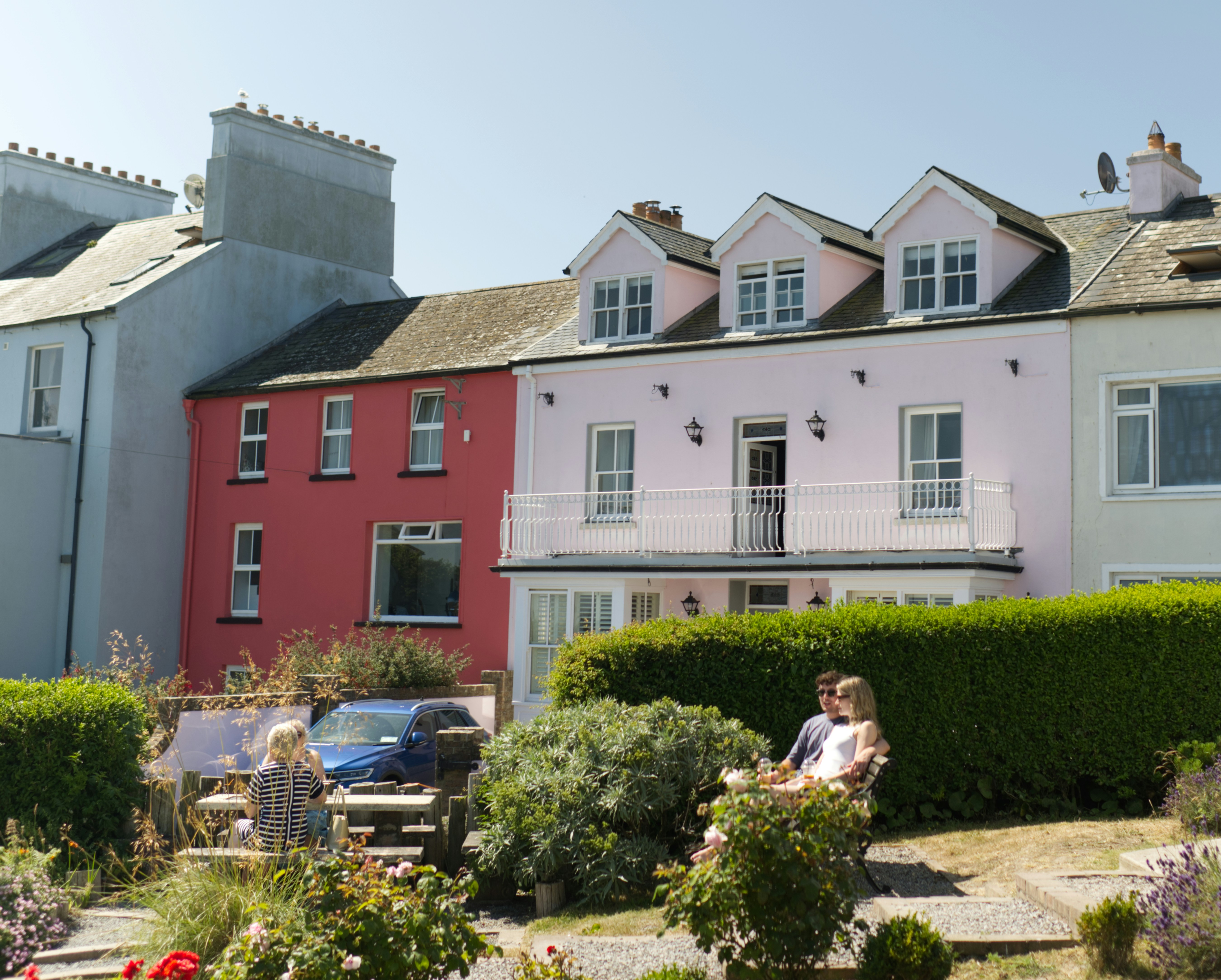 Colorful houses lined up along a garden, with a couple enjoying the sunny day in the foreground. The scene captures a serene coastal atmosphere.