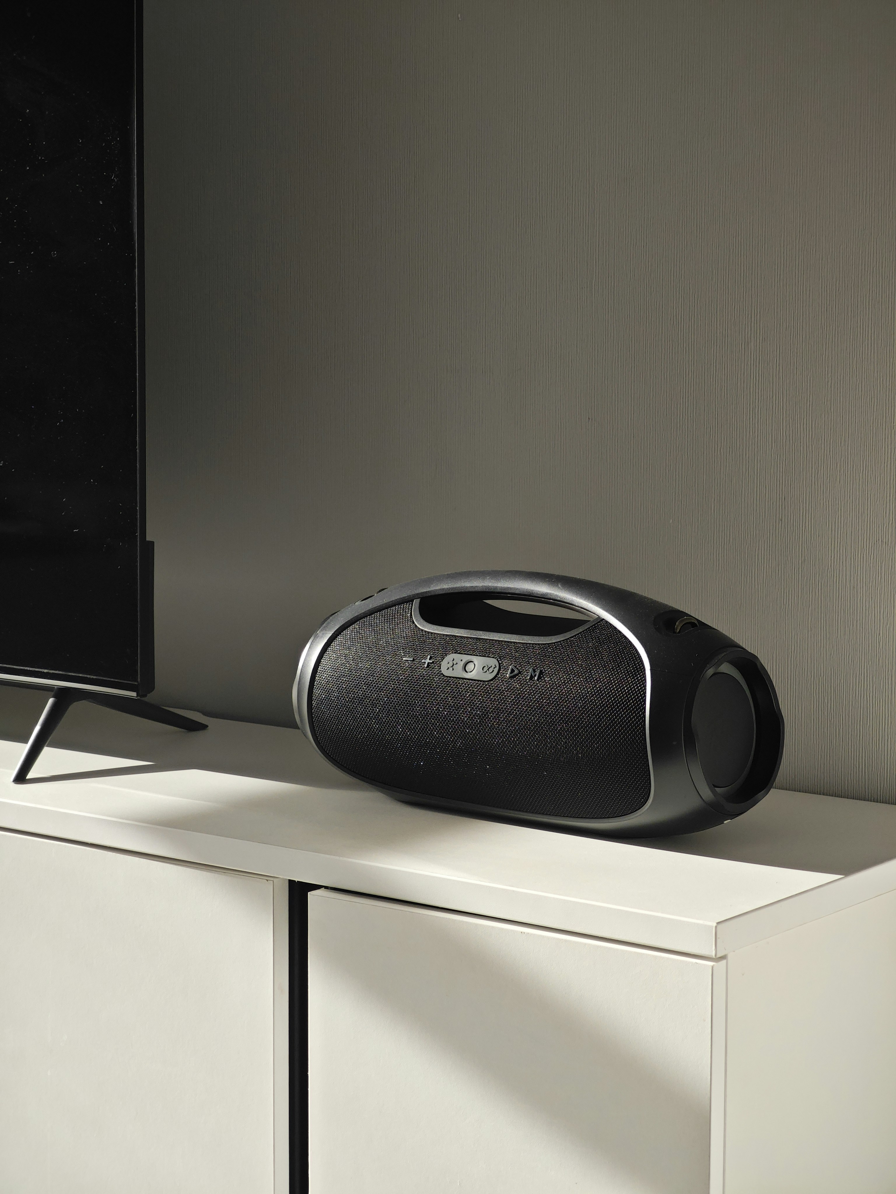 A sleek black speaker rests on a minimalist white surface, complemented by a dark television screen in the background. The soft lighting highlights the speaker's design and texture.