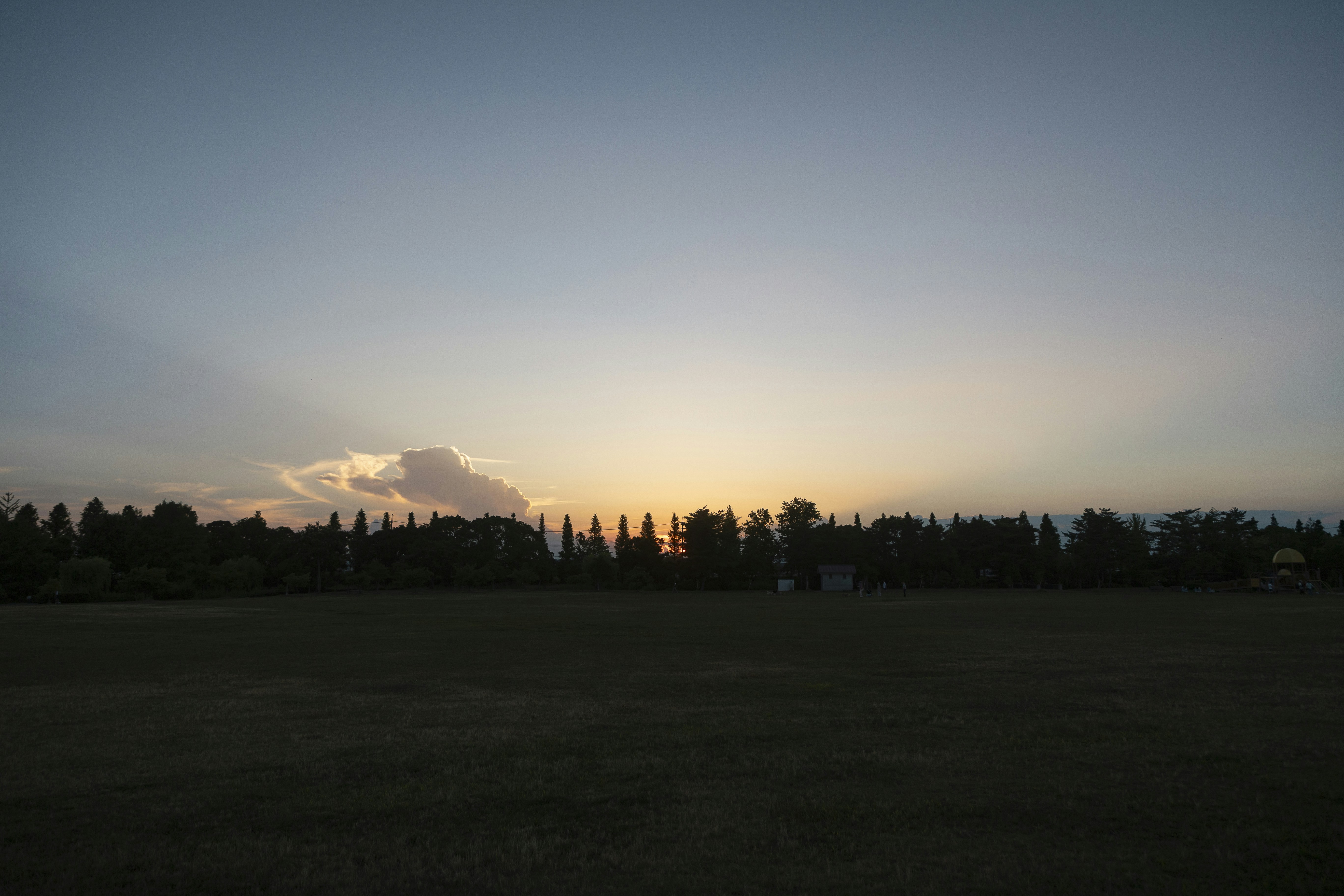 A serene twilight scene with a fading sun illuminating the sky, framed by silhouettes of trees on the horizon.