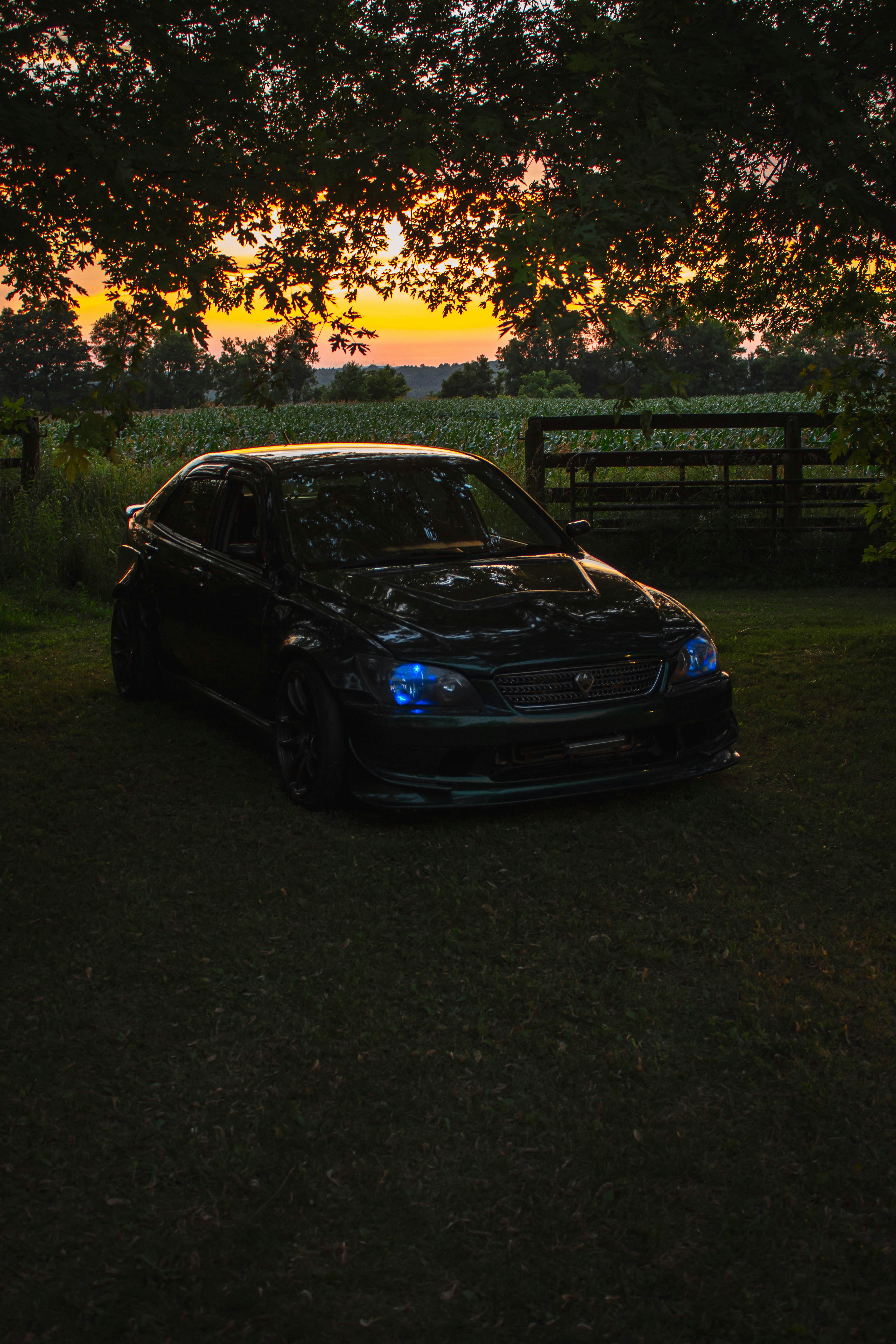 Modified car with glowing headlights parked on grass under a twilight sky, framed by trees and distant fields.