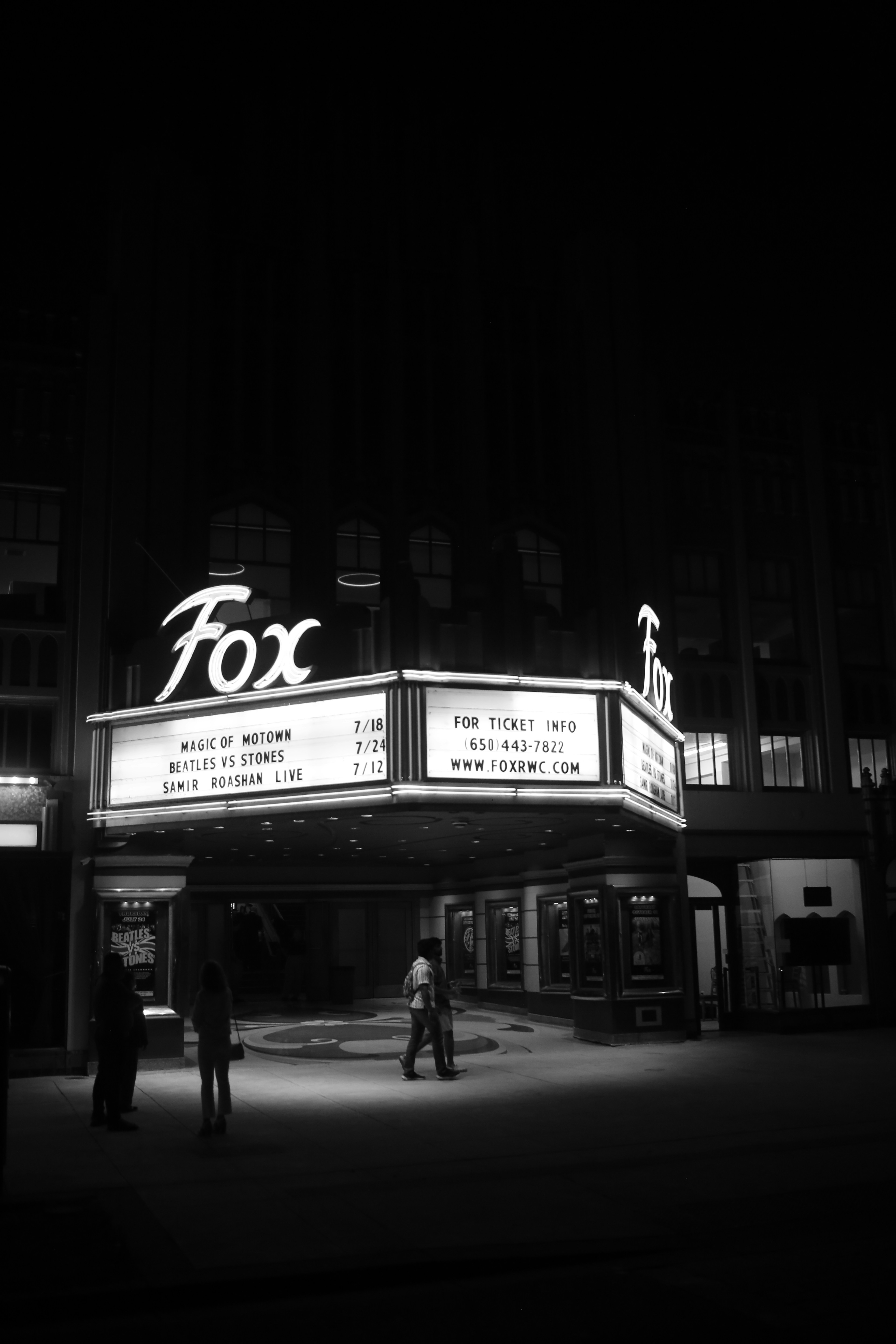 Illuminated marquee of the Fox Theater showcasing upcoming events, with silhouettes of pedestrians nearby.