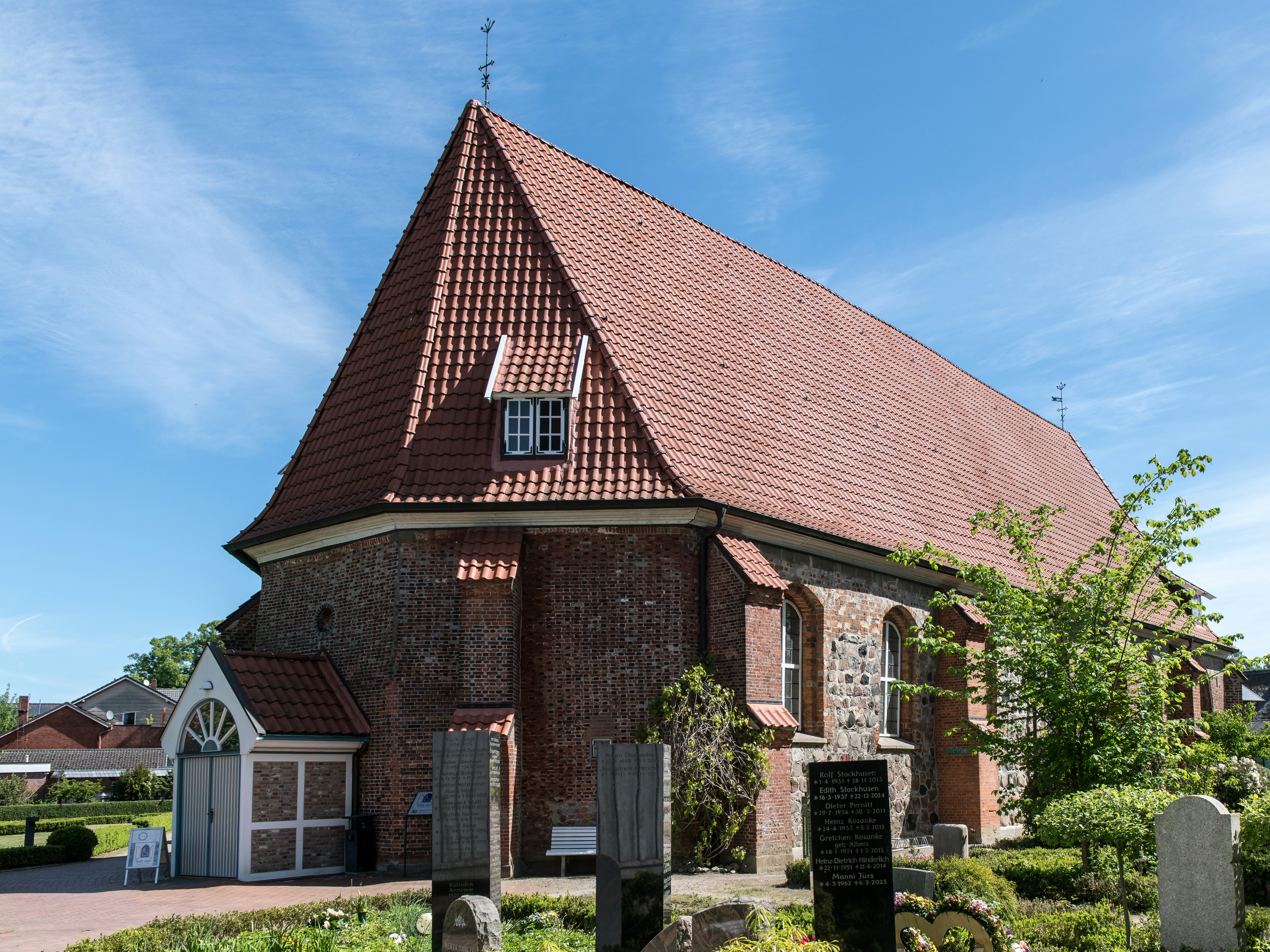 Historic brick church with a distinctive red roof, surrounded by lush greenery and gravestones, showcasing a blend of nature and history.