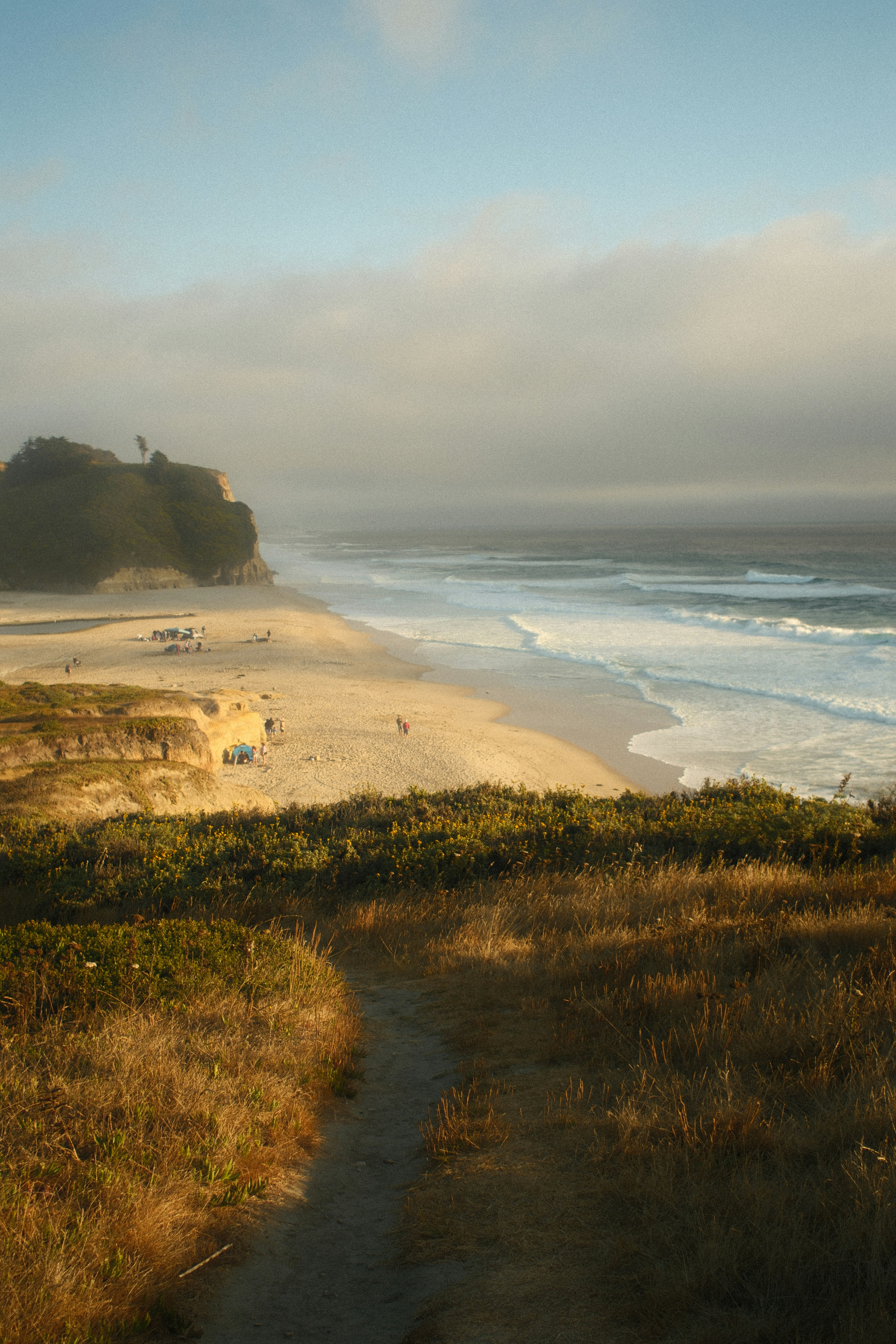 Golden beach at dusk with gentle waves lapping at the shore, framed by lush greenery and rocky cliffs. People enjoy the serene landscape.