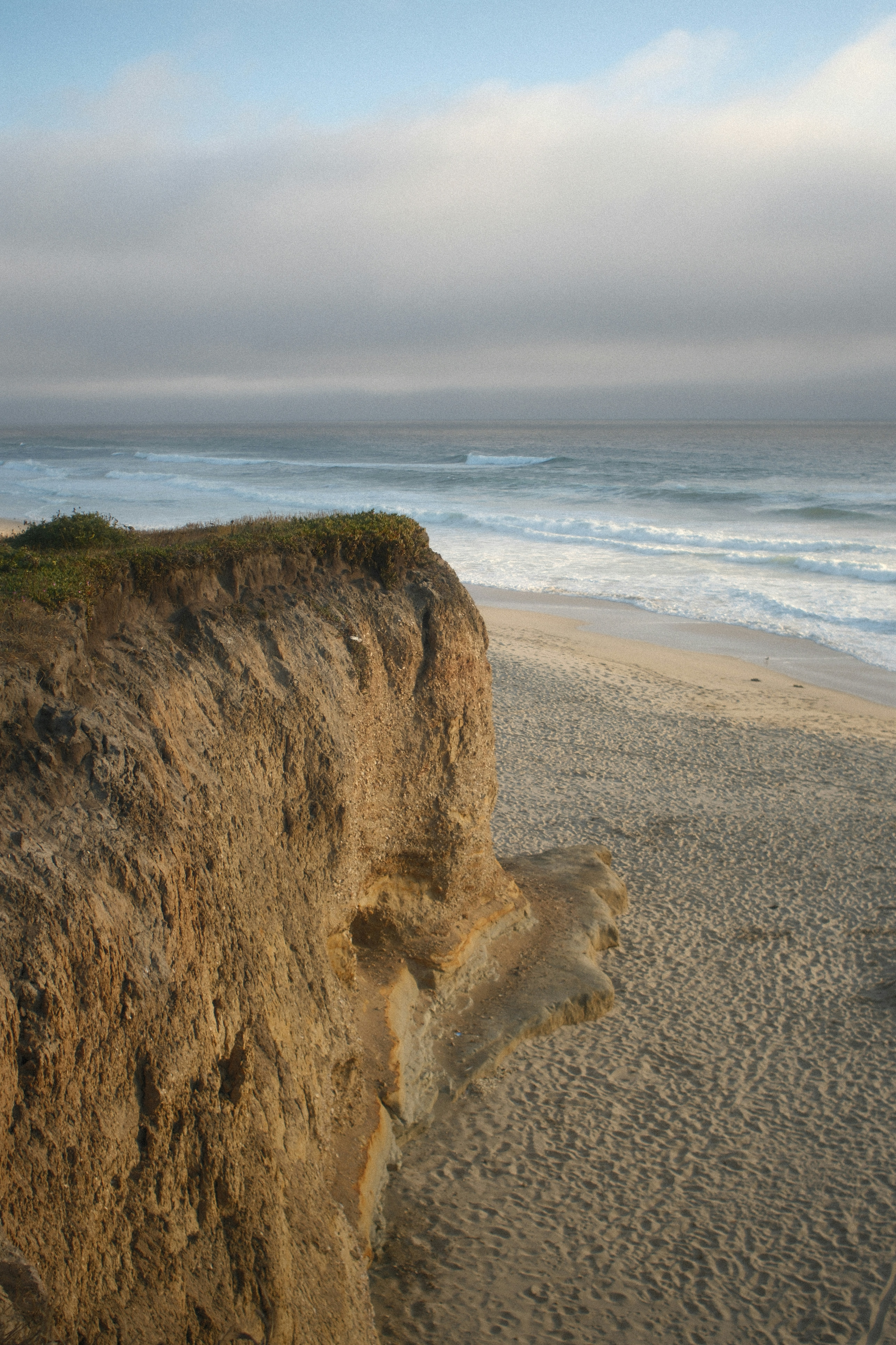 Rugged cliffside meeting the sandy shore, with gentle waves lapping at the base under a soft, overcast sky.