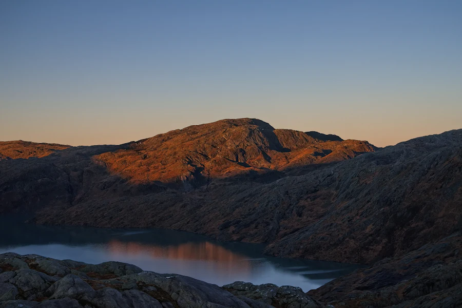 Sunlit rocky ridge glowing copper at golden hour with its reflection in a calm mountain tarn.
