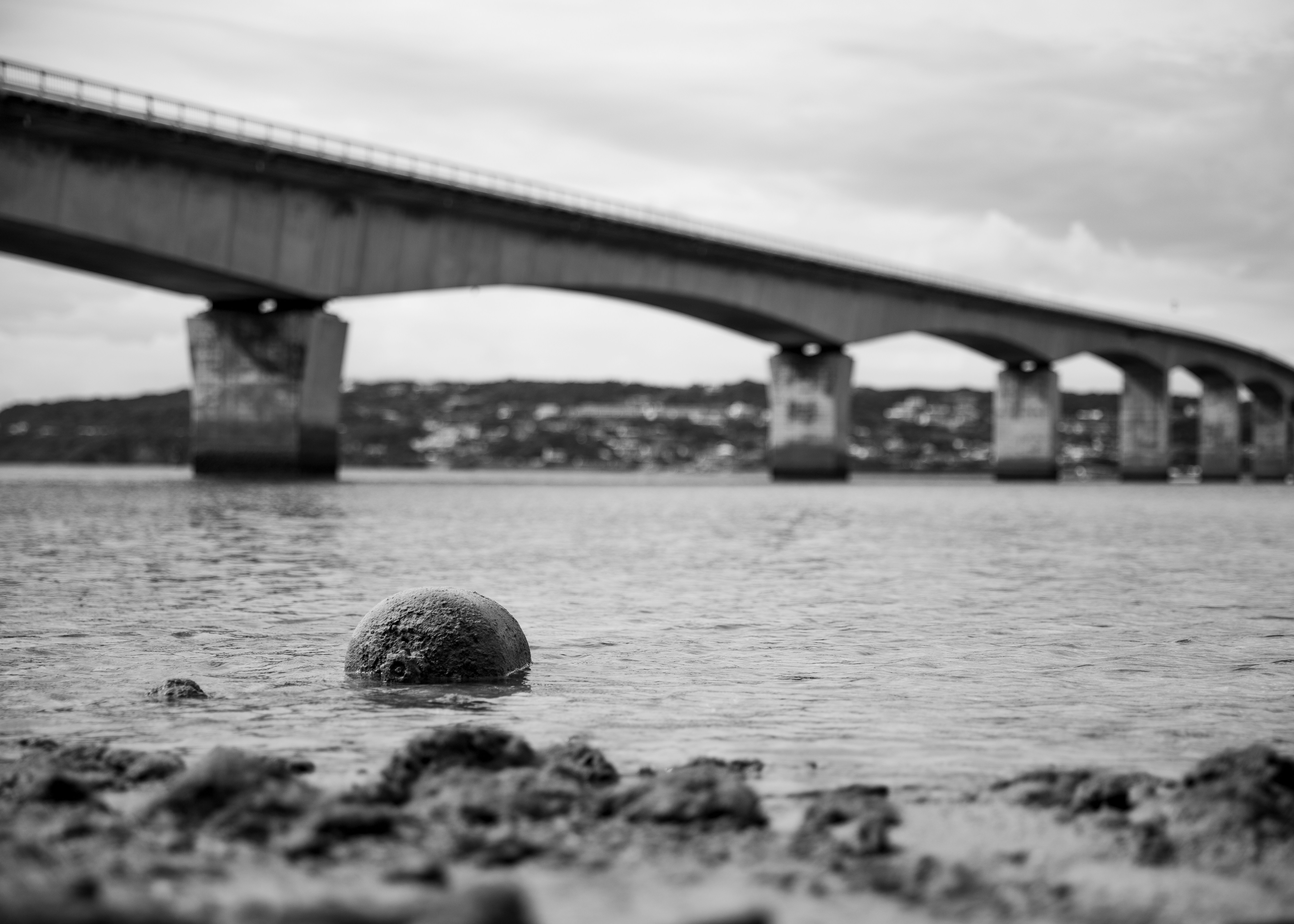 A weathered stone partially submerged in calm waters, with a bridge arching gracefully in the background. The monochrome palette adds a timeless quality to the scene.