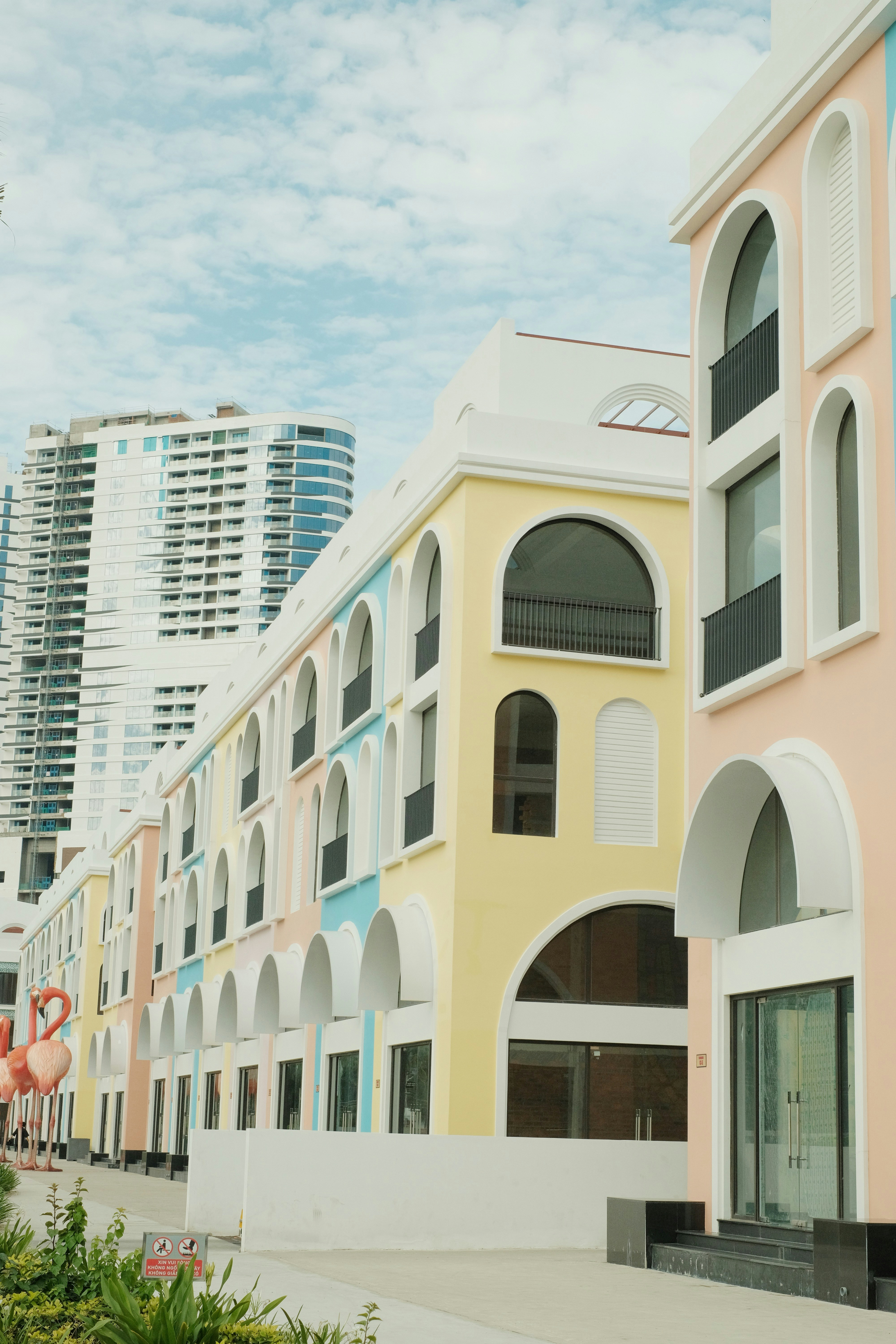 Colorful buildings with arched windows and a flamingo sculpture line the walkway, reflecting a blend of modern design and coastal charm.
