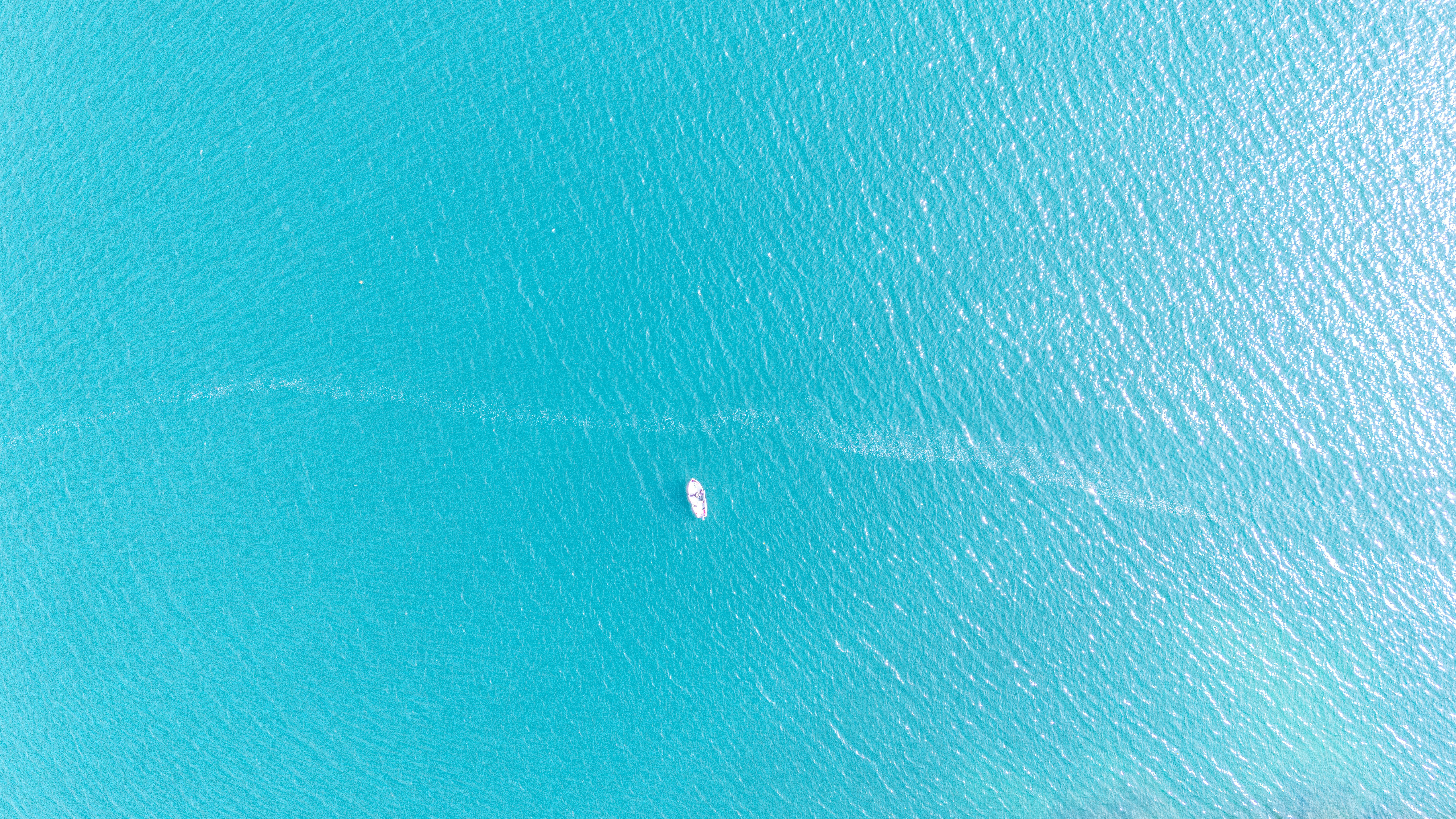 A lone boat glides over the tranquil turquoise waters, leaving a gentle wake behind. The vastness of the ocean creates a serene atmosphere.