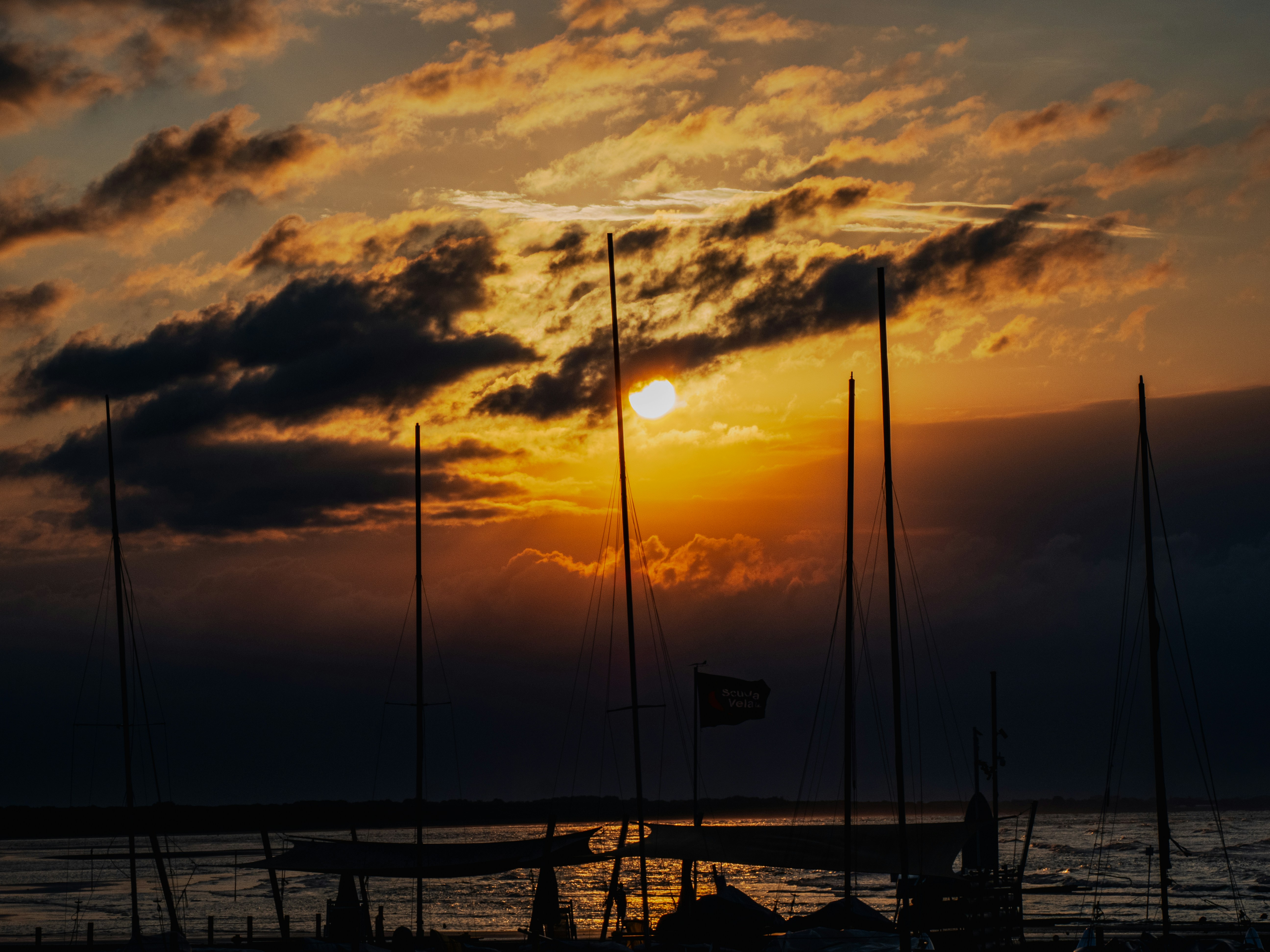 Sailboat masts silhouetted against a vibrant sunset, with clouds reflecting warm hues over the water.
