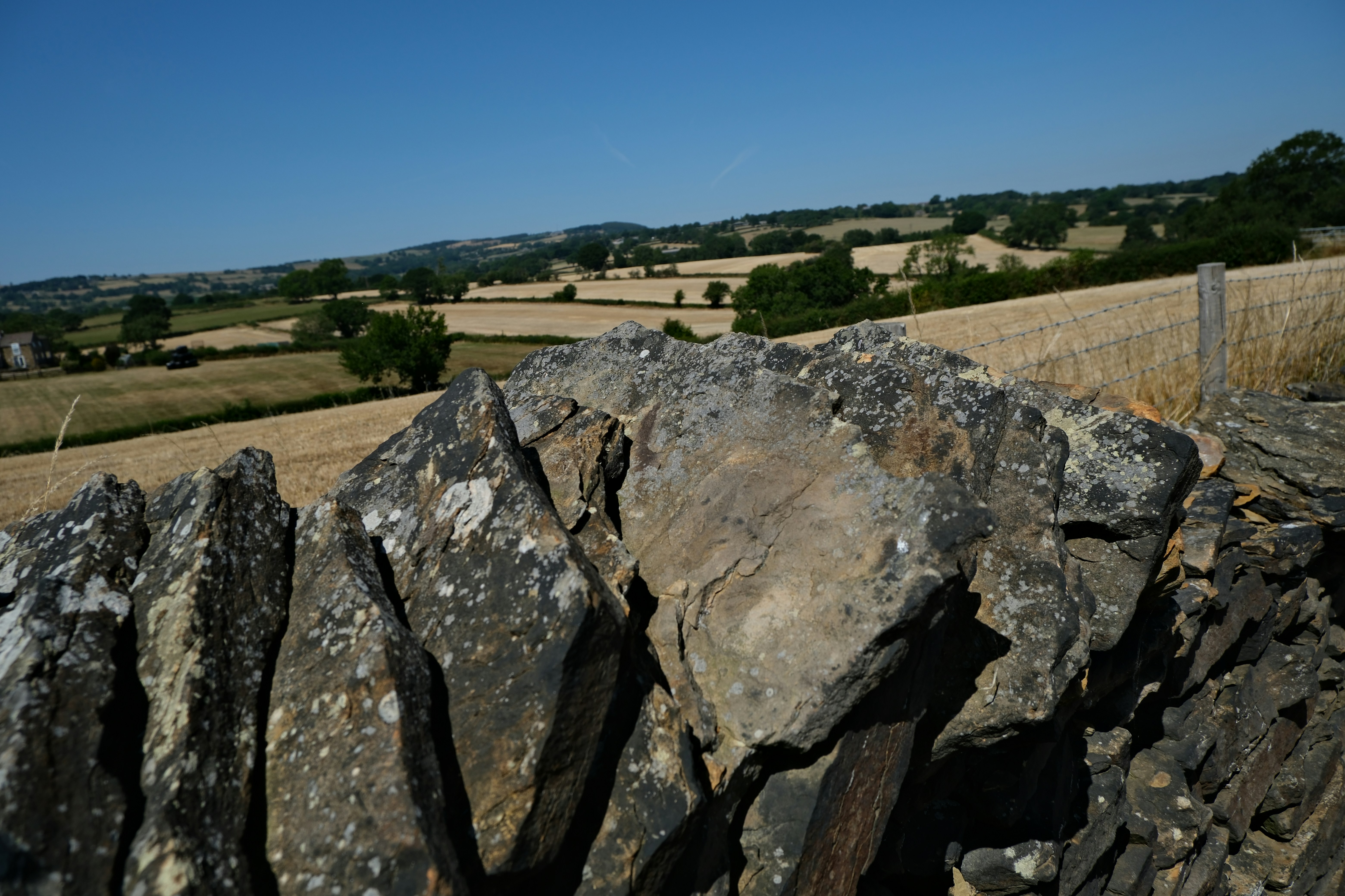 Close-up of a rugged stone wall with a panoramic view of golden fields and distant hills under a clear blue sky.