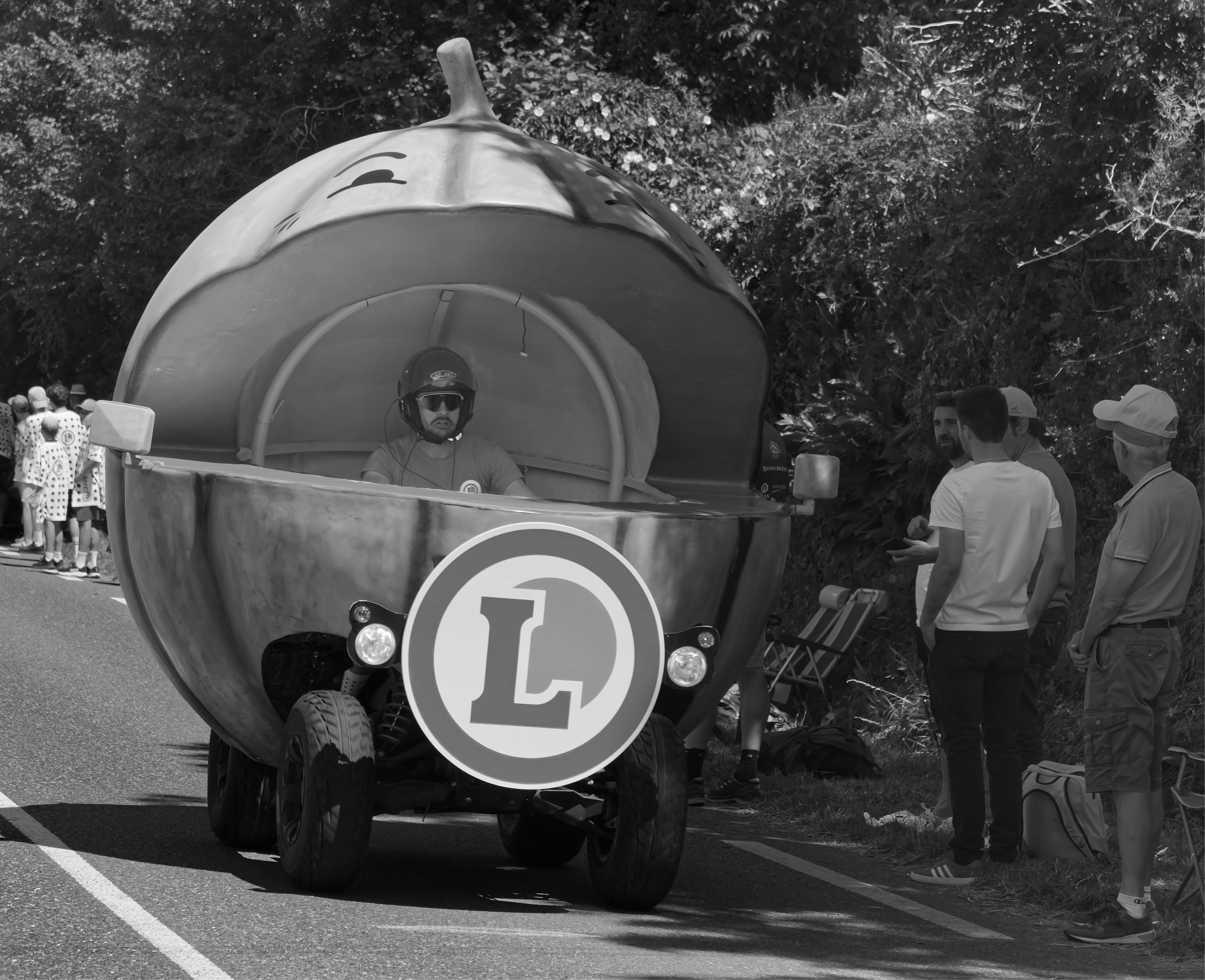 A whimsical vehicle shaped like a giant acorn drives down a road, capturing the attention of onlookers at a parade.