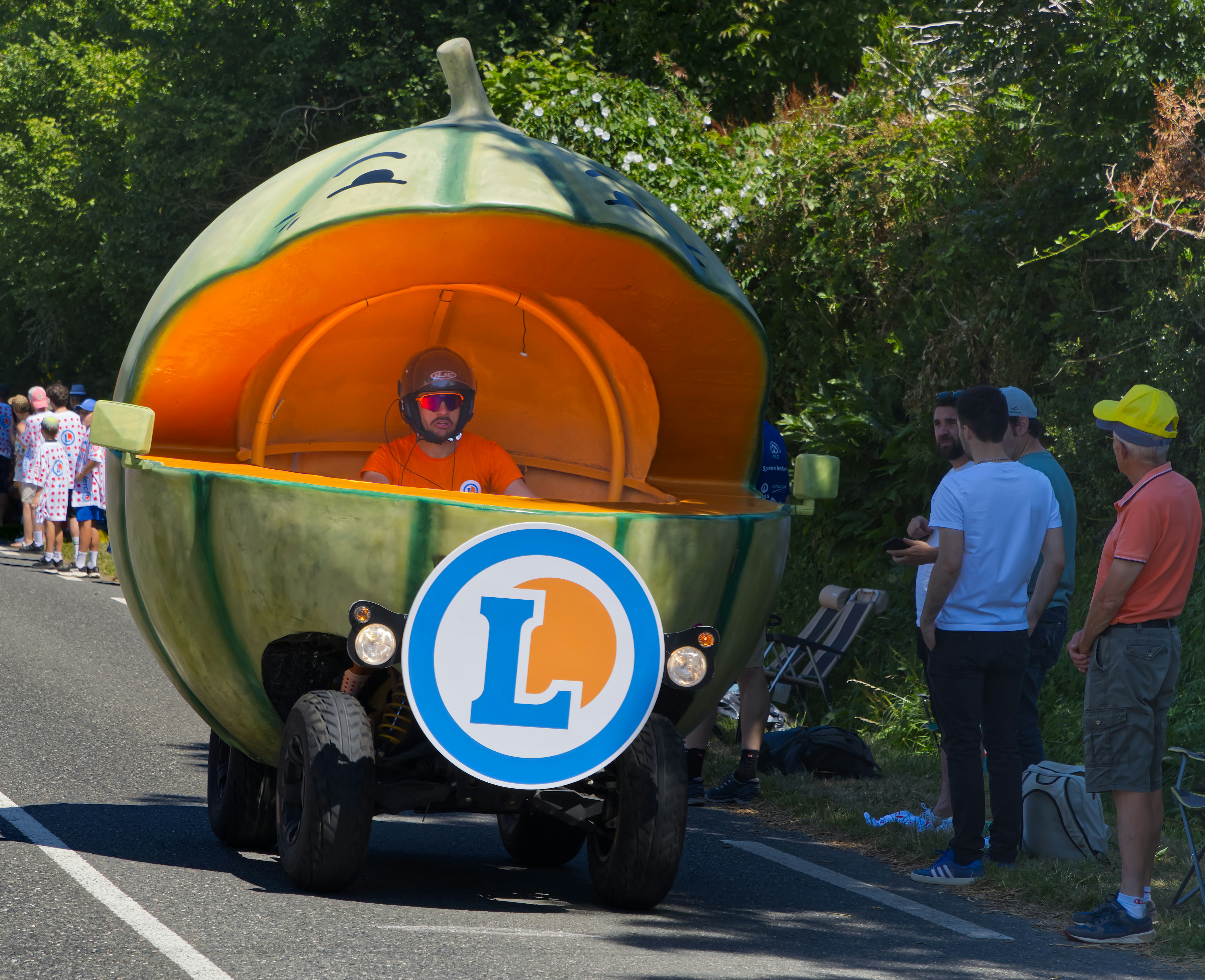 Tour de France 2025 : 7ème étape de Saint-Malo à Mur-de-Bretagne. Un des sponsors de la caravane du Tour a pris le melon! | A man rides in a melon car on a road.