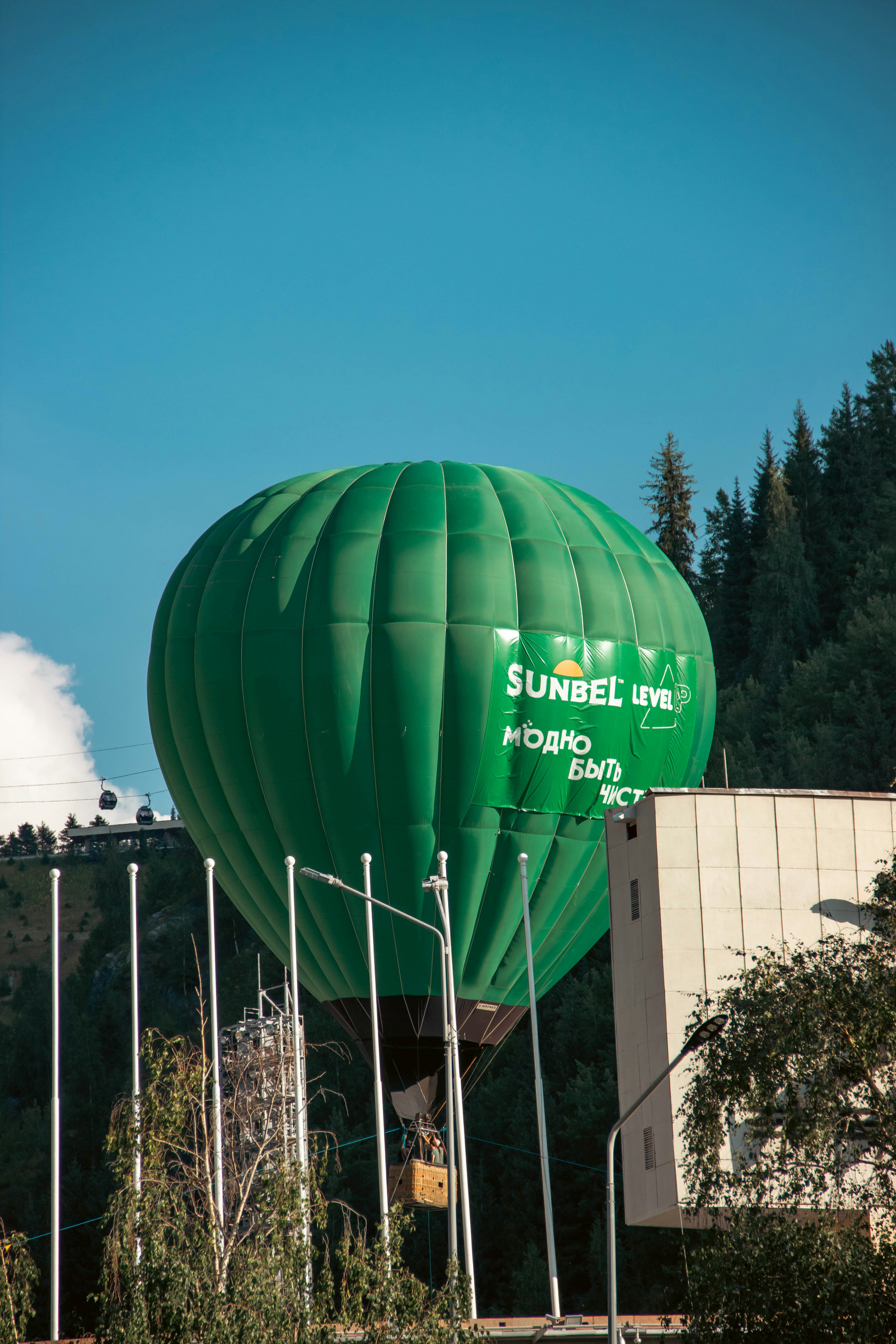 A vibrant green hot air balloon emblazoned with a logo, hovering above a building amidst a backdrop of trees and blue sky.
