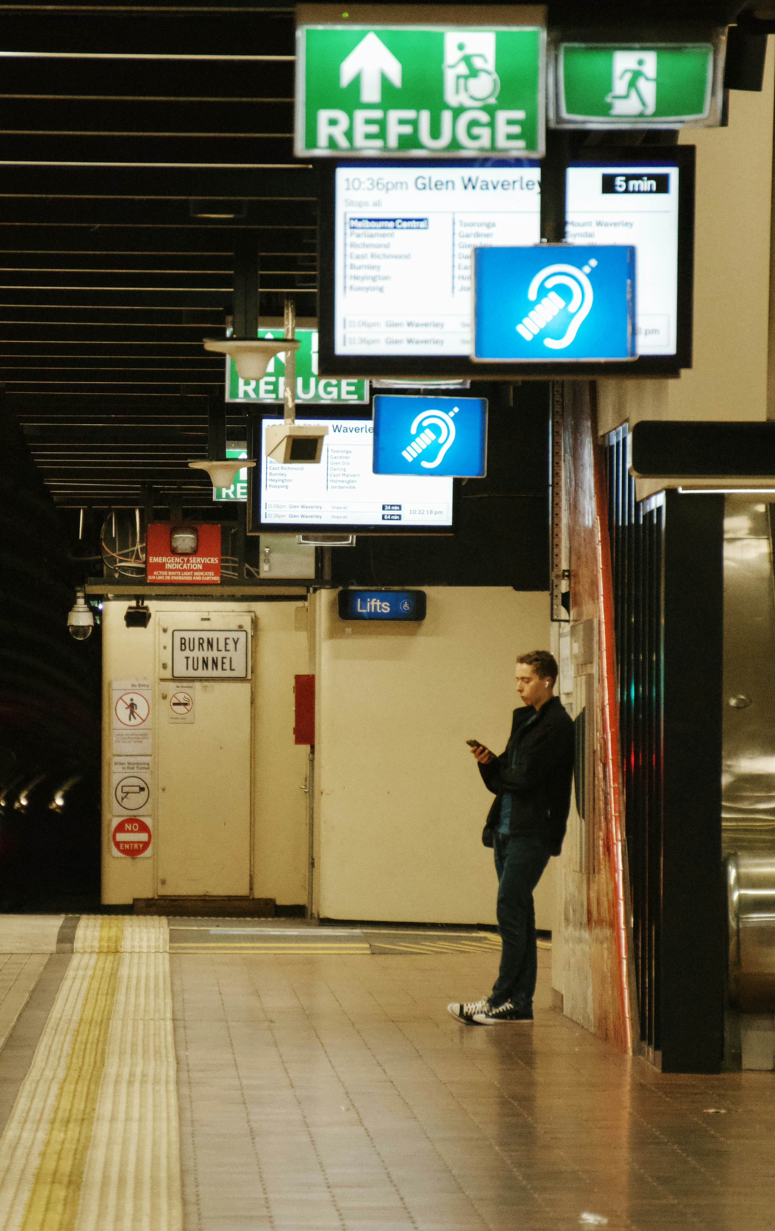 A lone figure stands on a subway platform, engrossed in their phone, surrounded by illuminated signs and directional indicators.