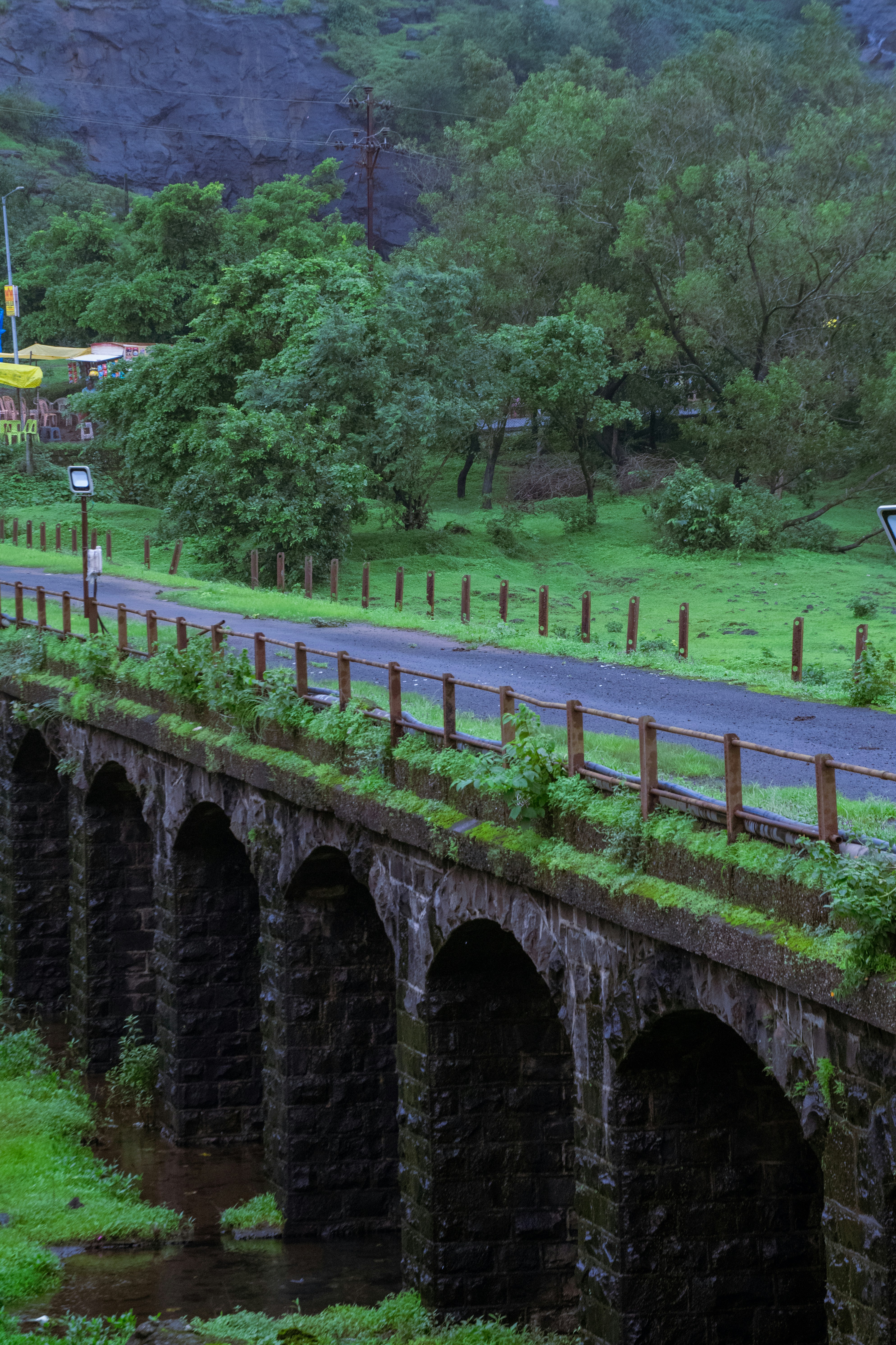 Historic stone viaduct spanning a tranquil stream, surrounded by vibrant greenery and a winding road. 