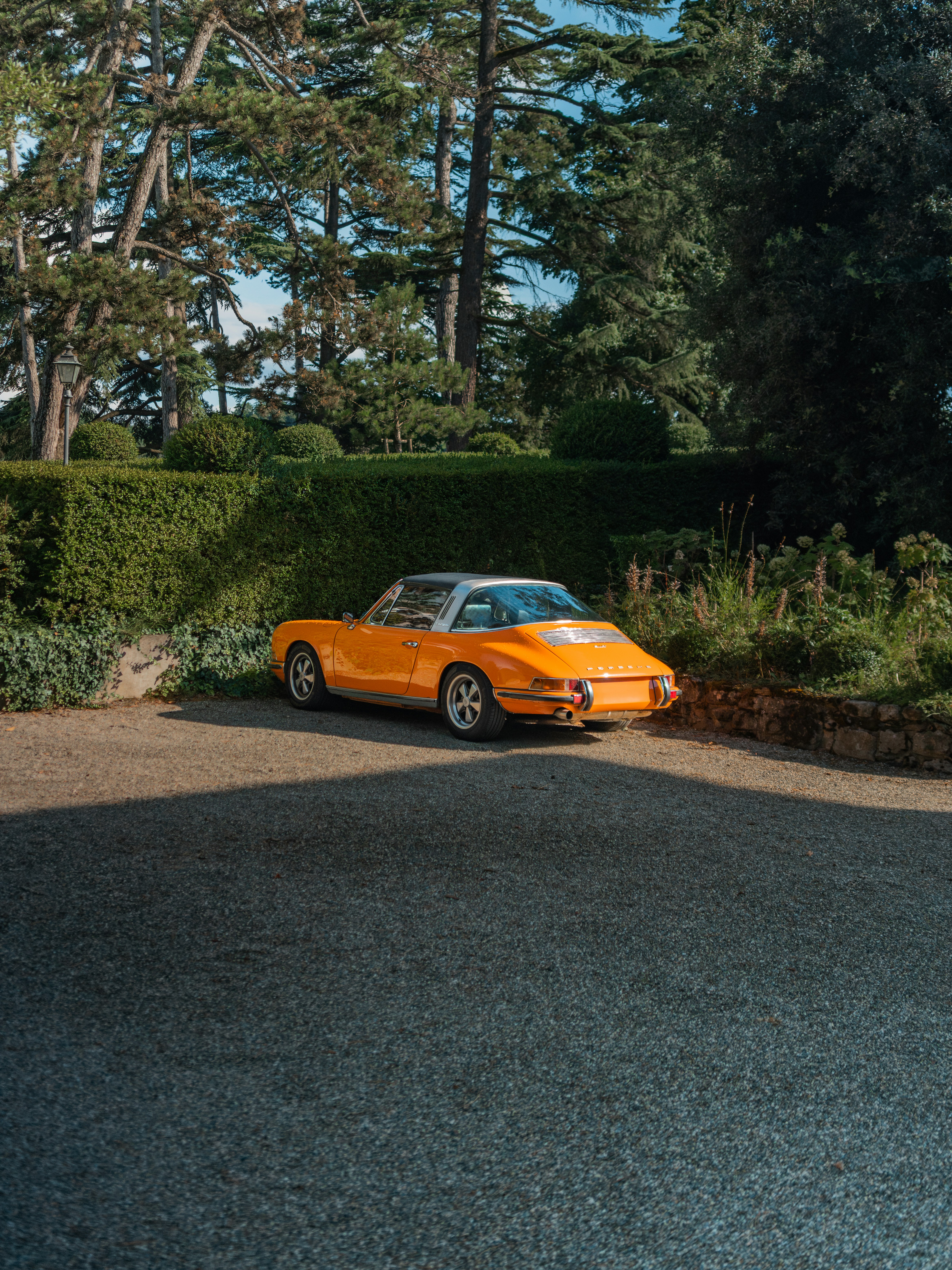 An orange classic car parked beside a neatly trimmed hedge and surrounded by greenery. The scene captures a serene moment in a garden setting.