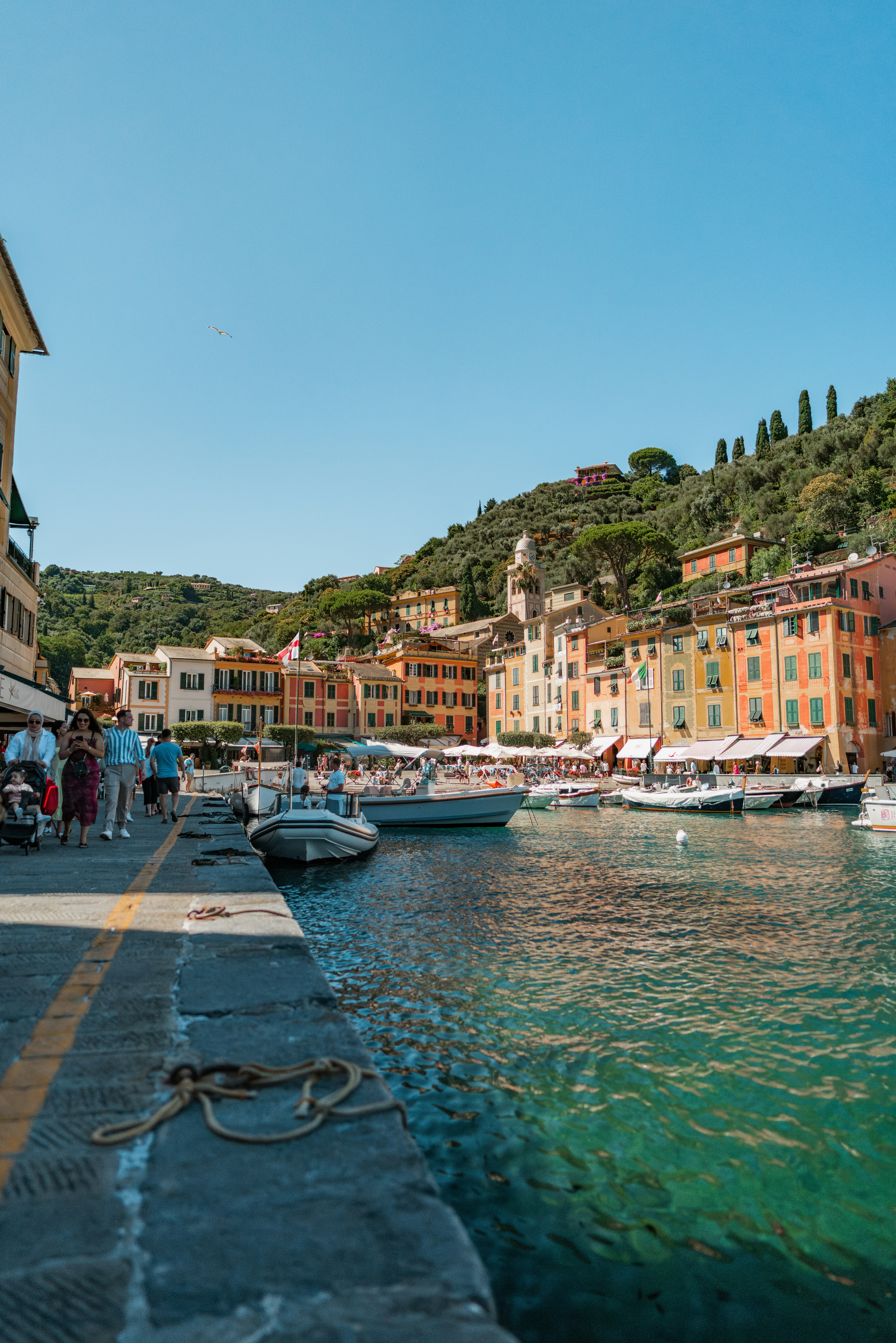 Charming waterfront scene in Portofino, Italy, featuring vibrant buildings, boats, and a bustling promenade. The lush hillside adds a picturesque backdrop.