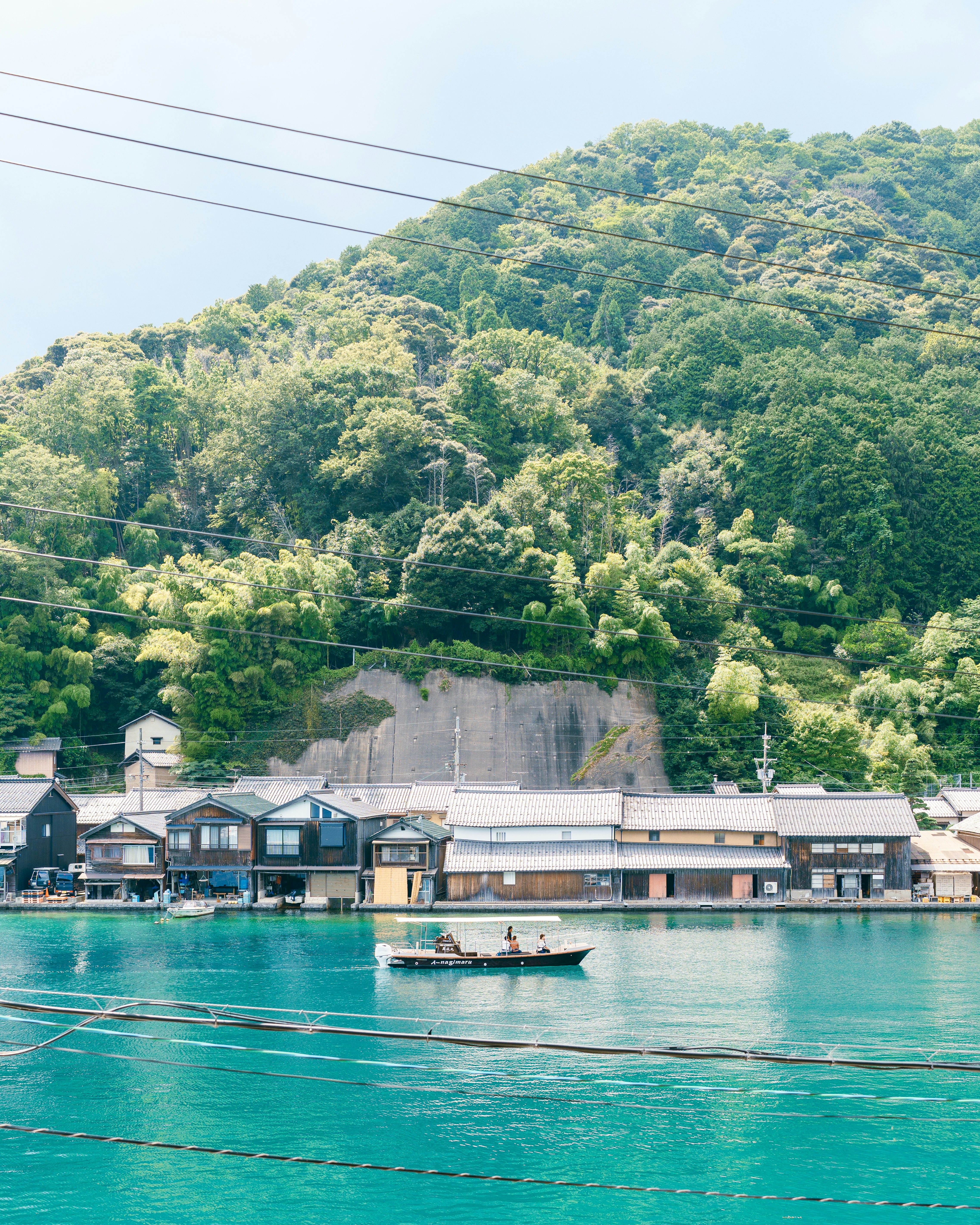 A tranquil scene featuring traditional houses lining a serene waterfront, with a small boat carrying passengers gliding through the turquoise waters.