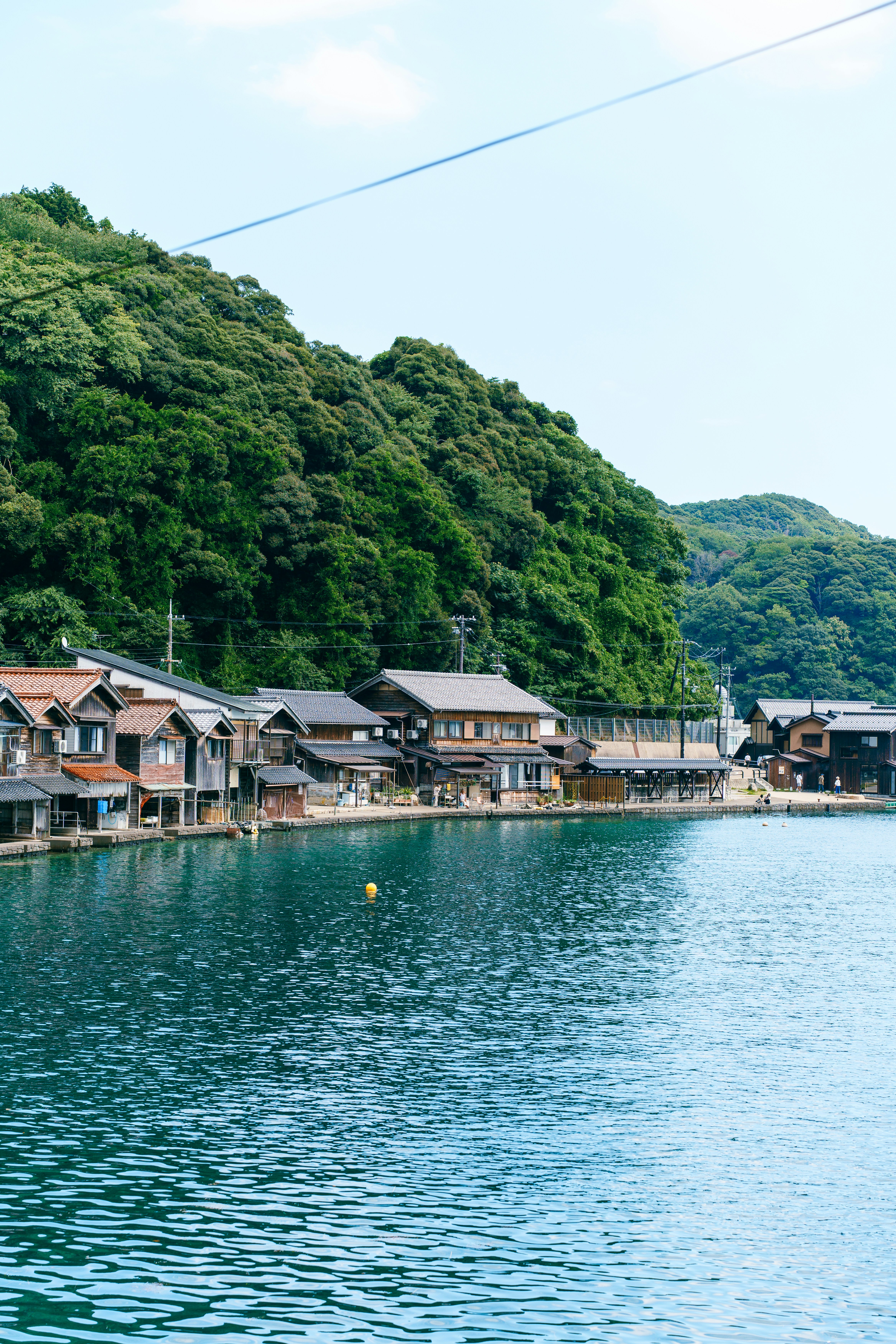 Quaint wooden houses line the tranquil waters of a coastal village, framed by lush green hills. The scene captures the essence of peaceful seaside living.