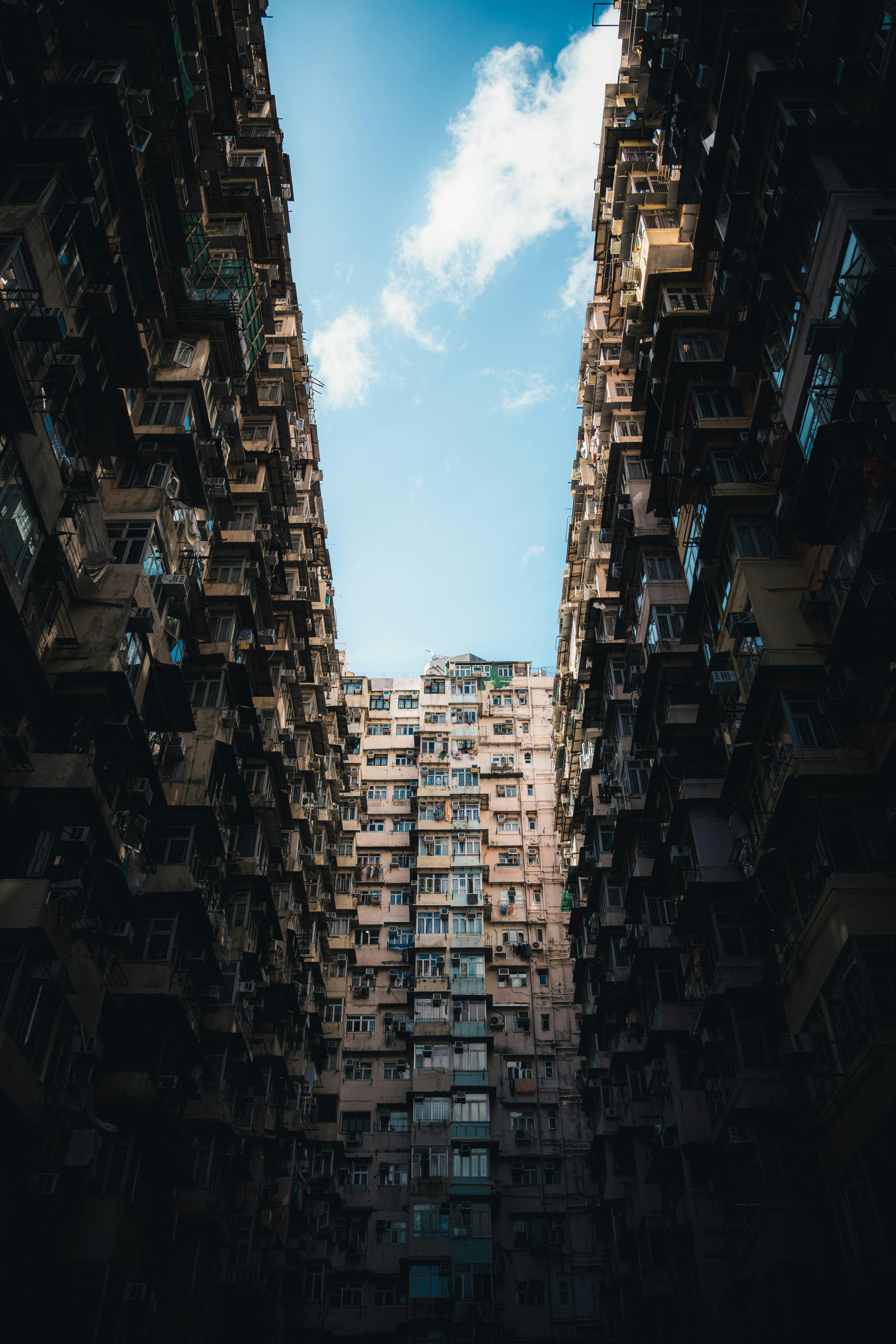 High-rise buildings create a narrow passage, revealing a patch of blue sky above. The architectural density emphasizes urban life.