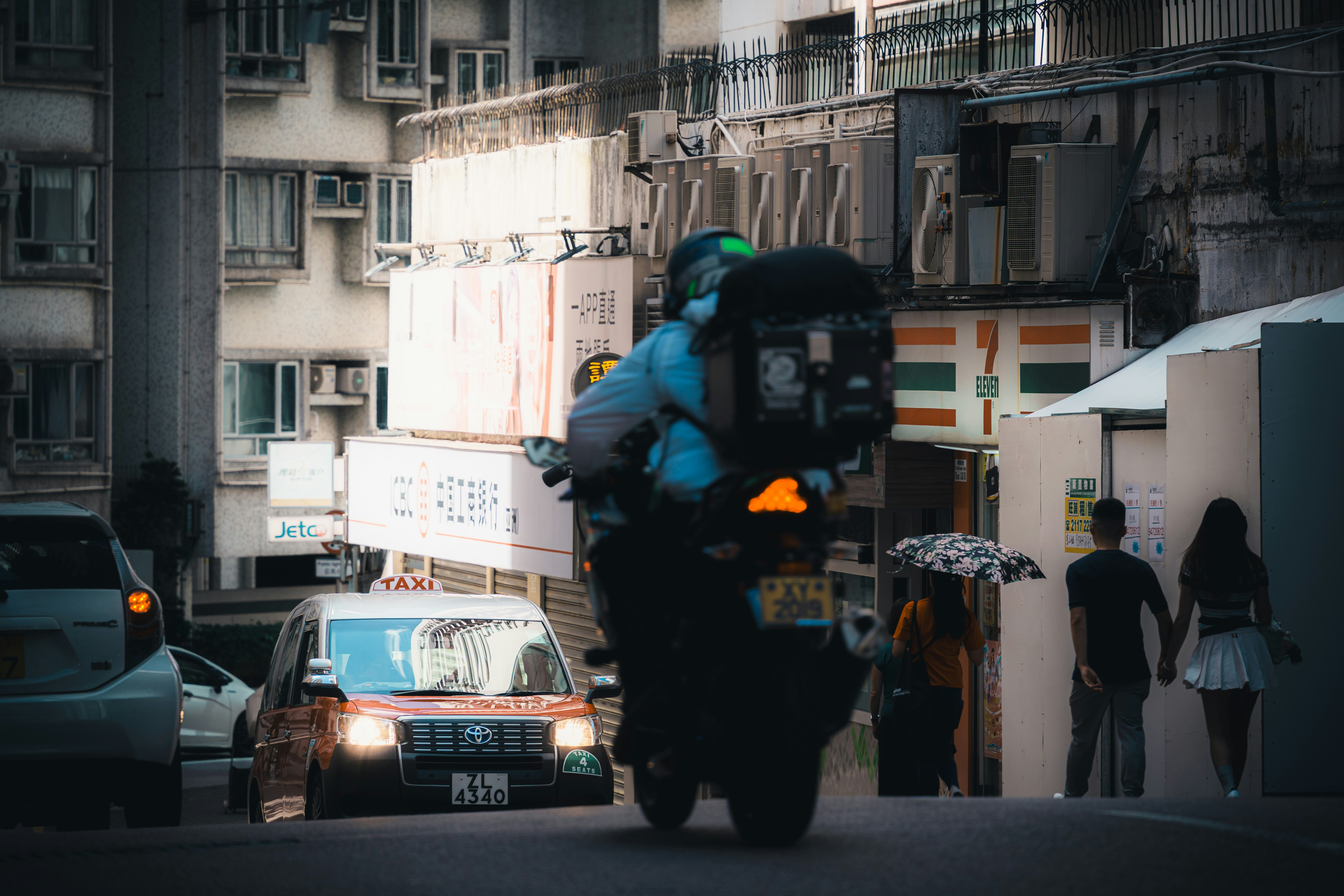 Motorcyclist navigating a bustling street with a taxi and pedestrians in the background. The scene captures the vibrancy of urban life.