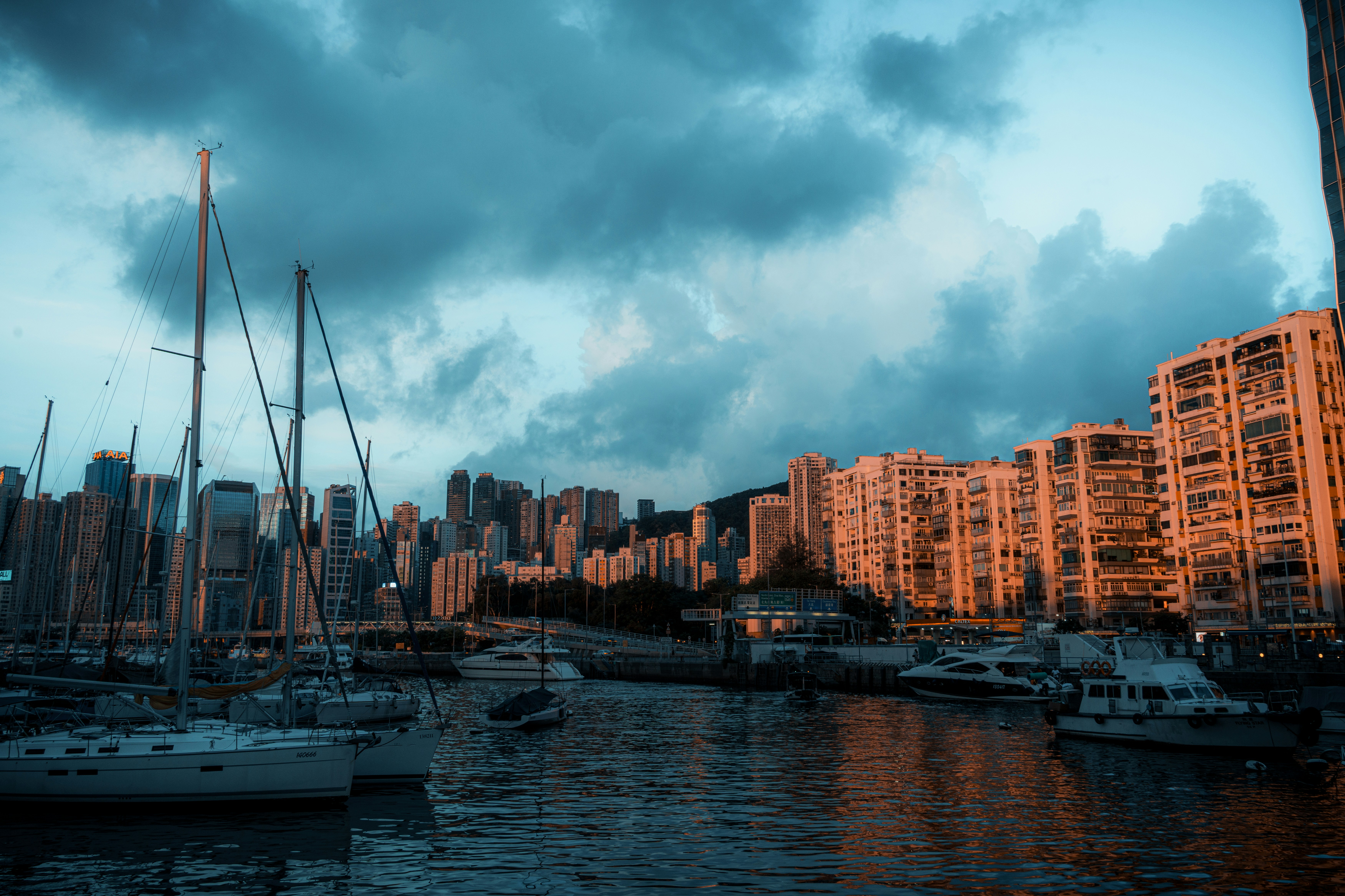 Sailboats docked in a vibrant harbor with city skyscrapers illuminated by the evening light and dramatic clouds overhead.