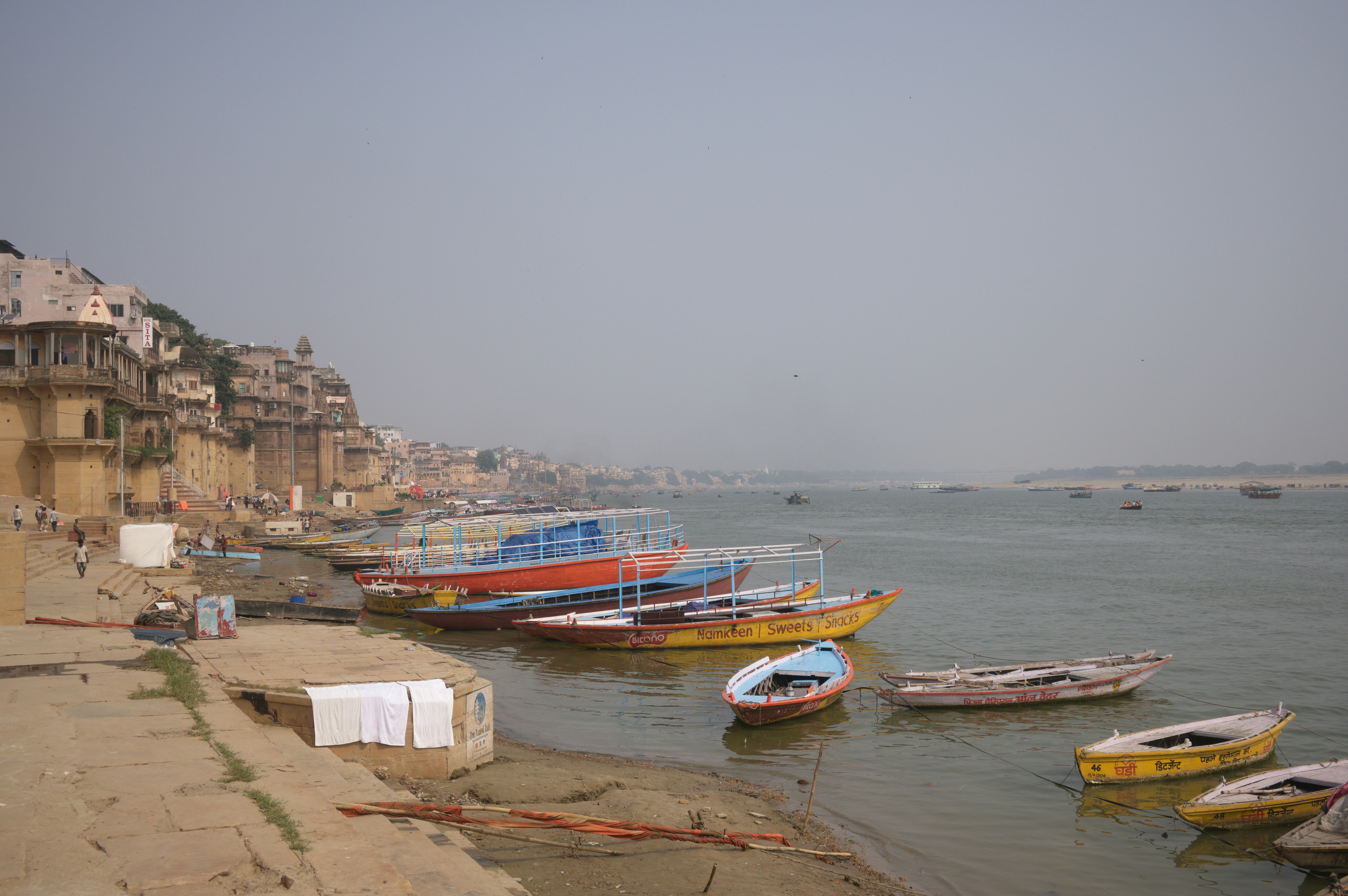 Colorful boats resting along the banks of the Ganges River, with historic buildings lining the shore under a hazy sky.
