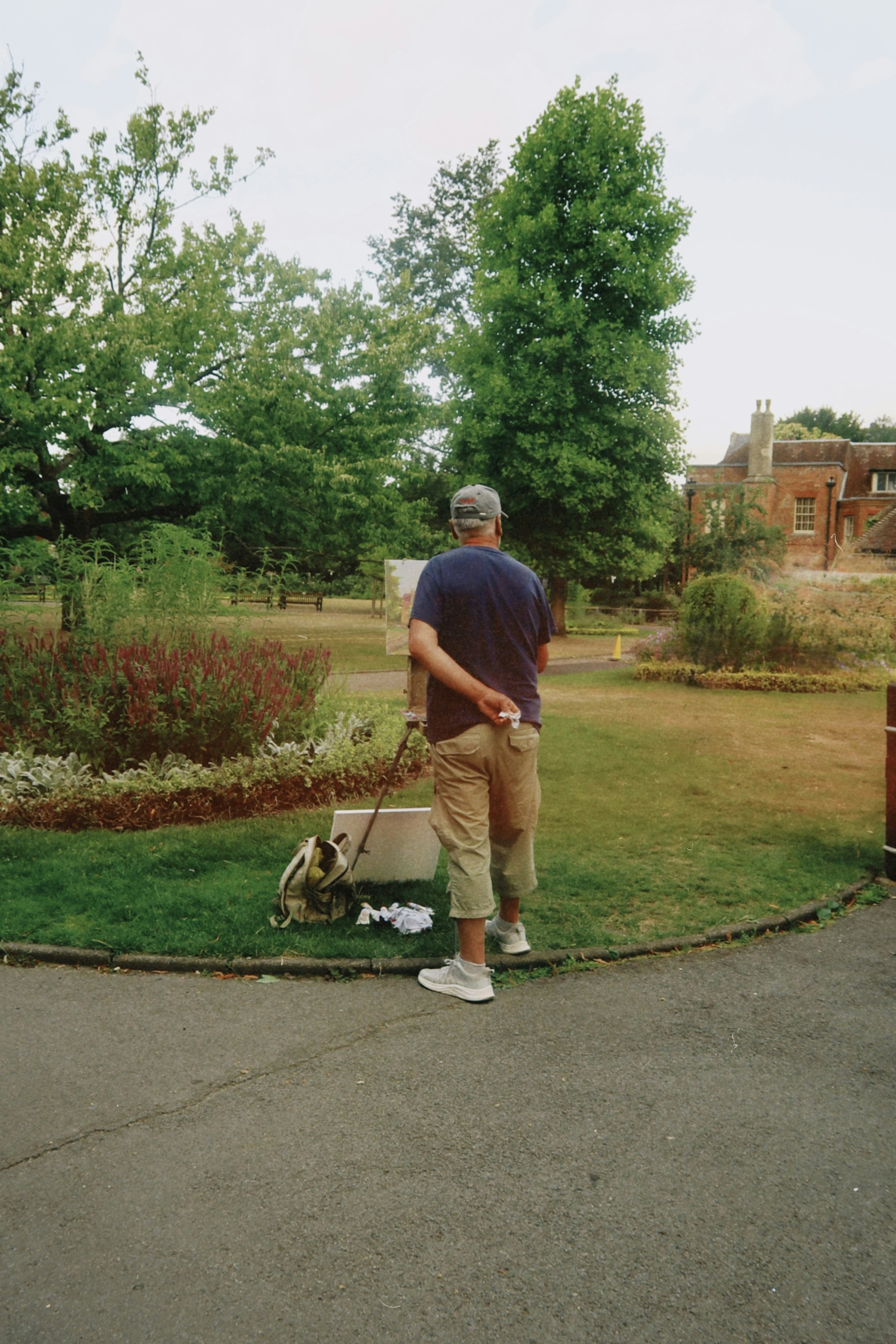 Man standing in a garden with a dog, observing the surroundings amid lush greenery and a historic building in the background.