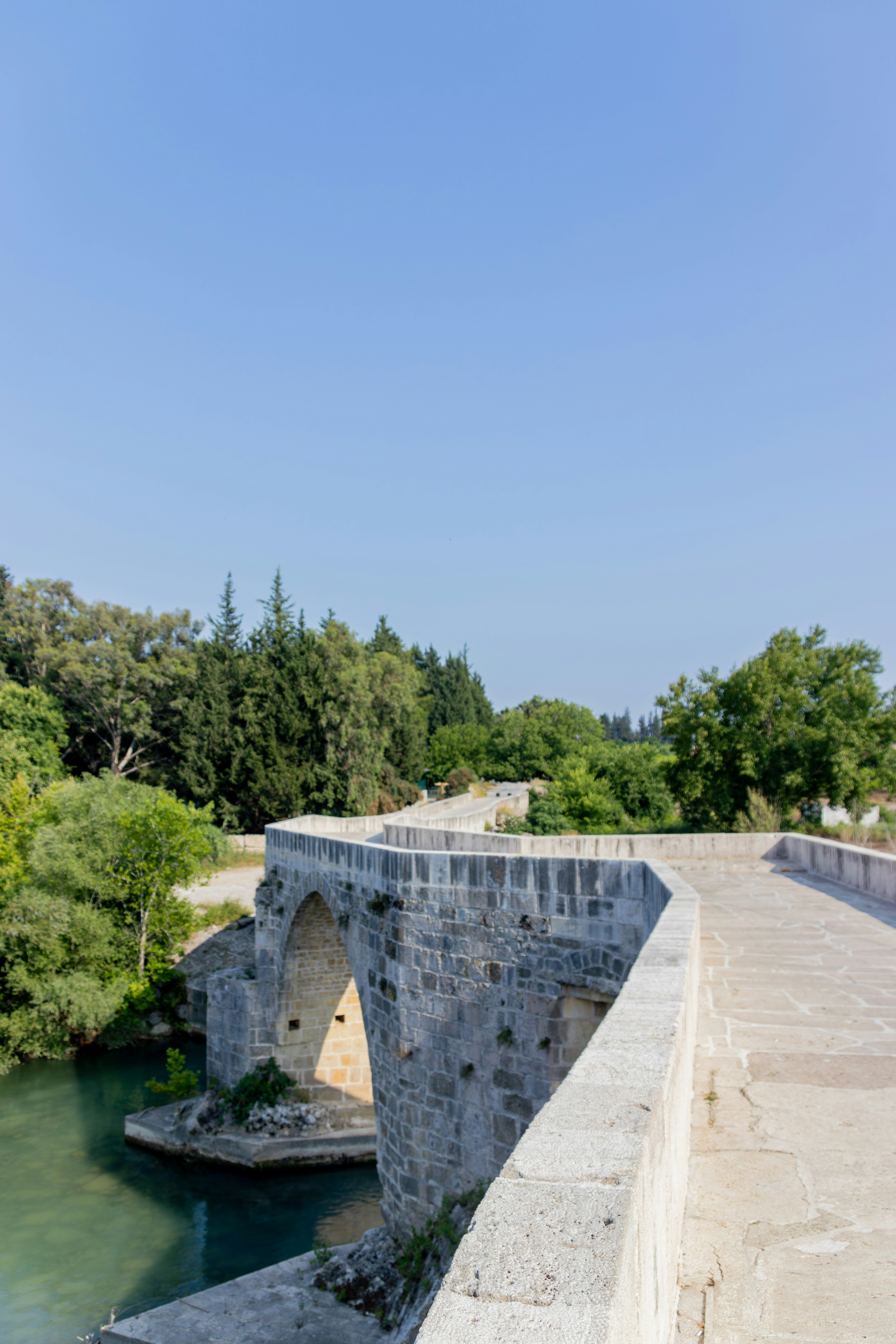 Historic stone bridge arching over a serene river, flanked by lush greenery under a clear blue sky.