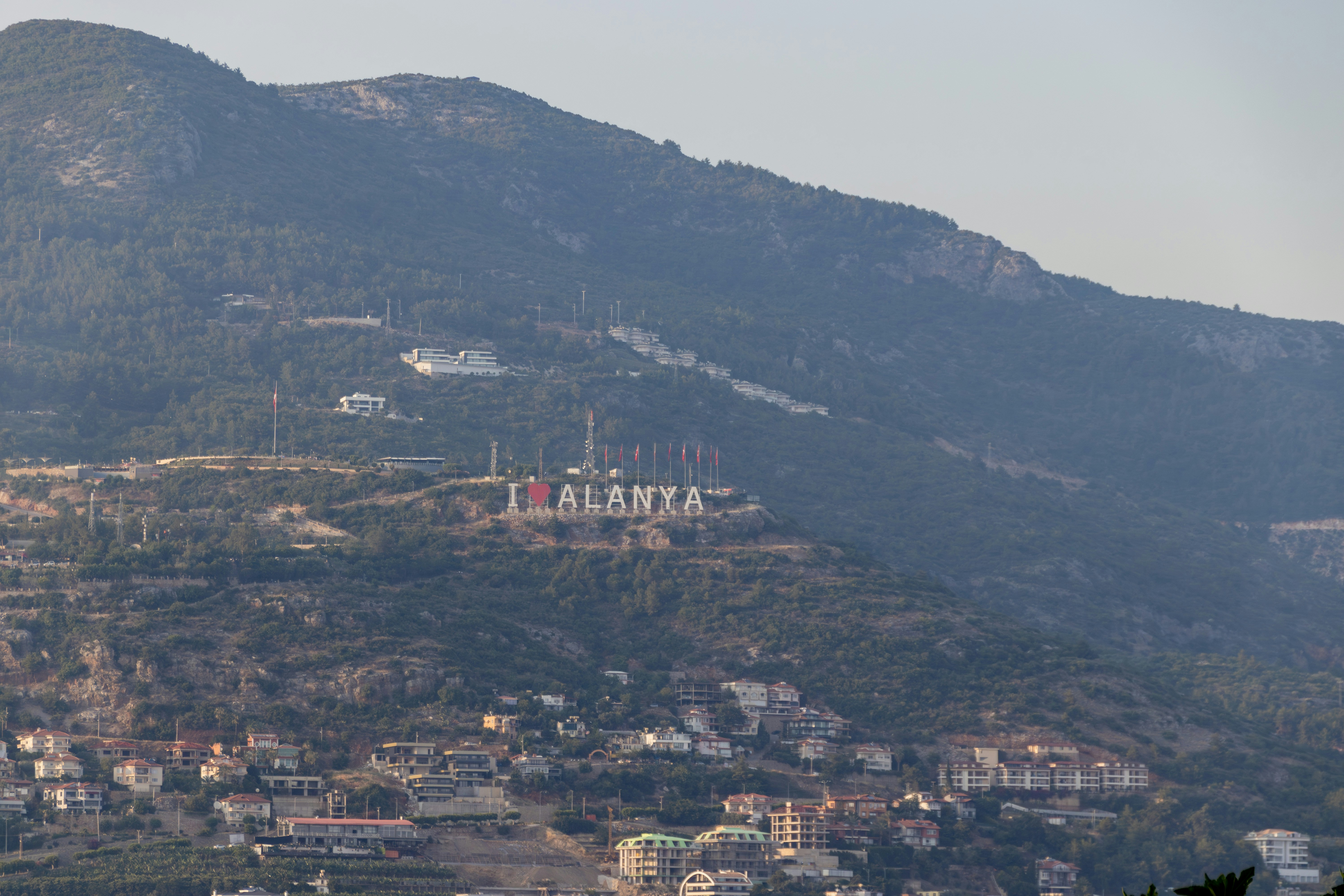 The iconic Alanya sign perched on a hillside, surrounded by lush greenery and residential buildings below.