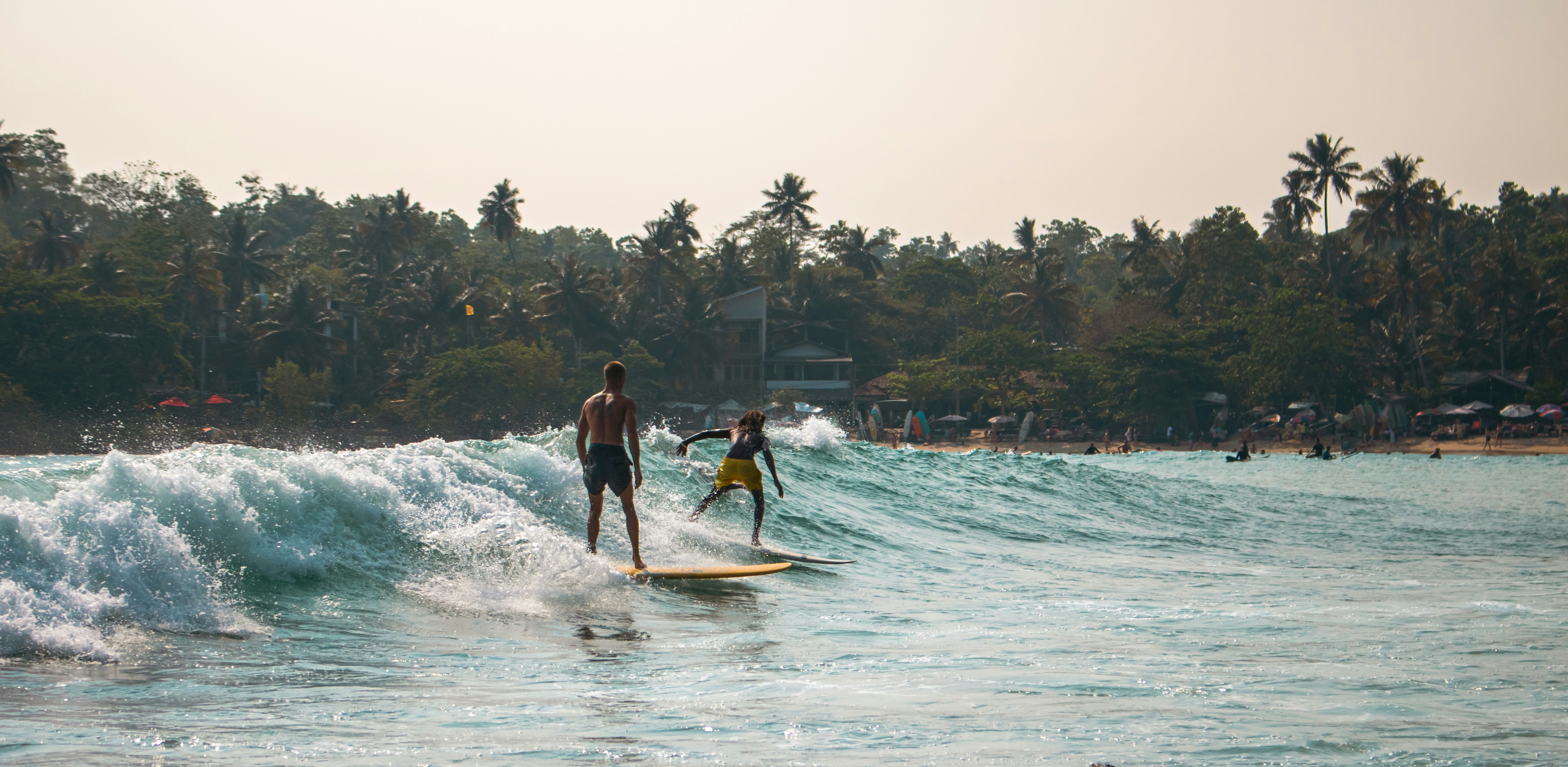 Two surfers skillfully riding waves under a warm sun, with palm trees and beachgoers in the background.