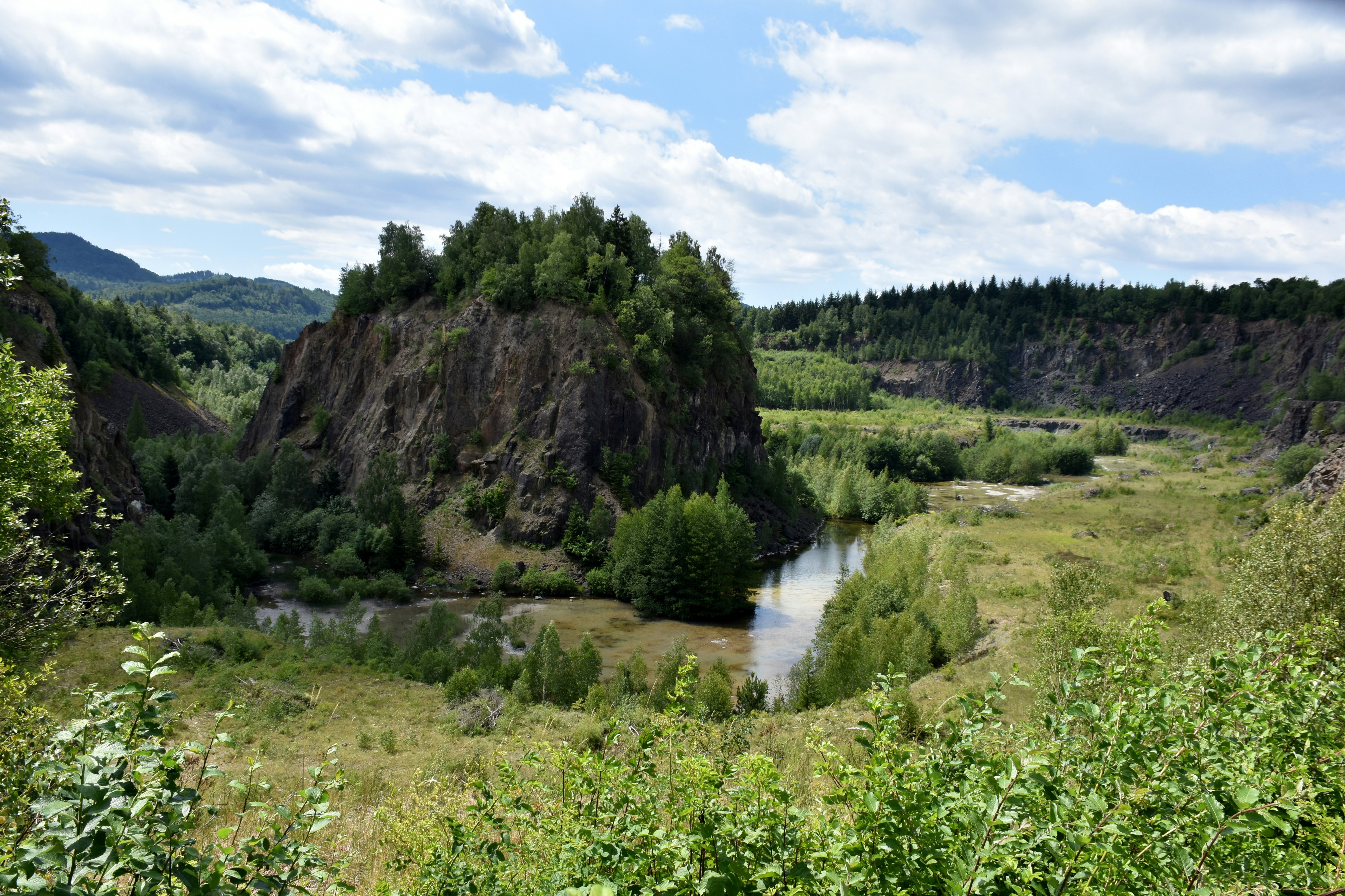 Lush green valley with a winding river surrounded by rocky outcrops and dense trees under a bright blue sky.