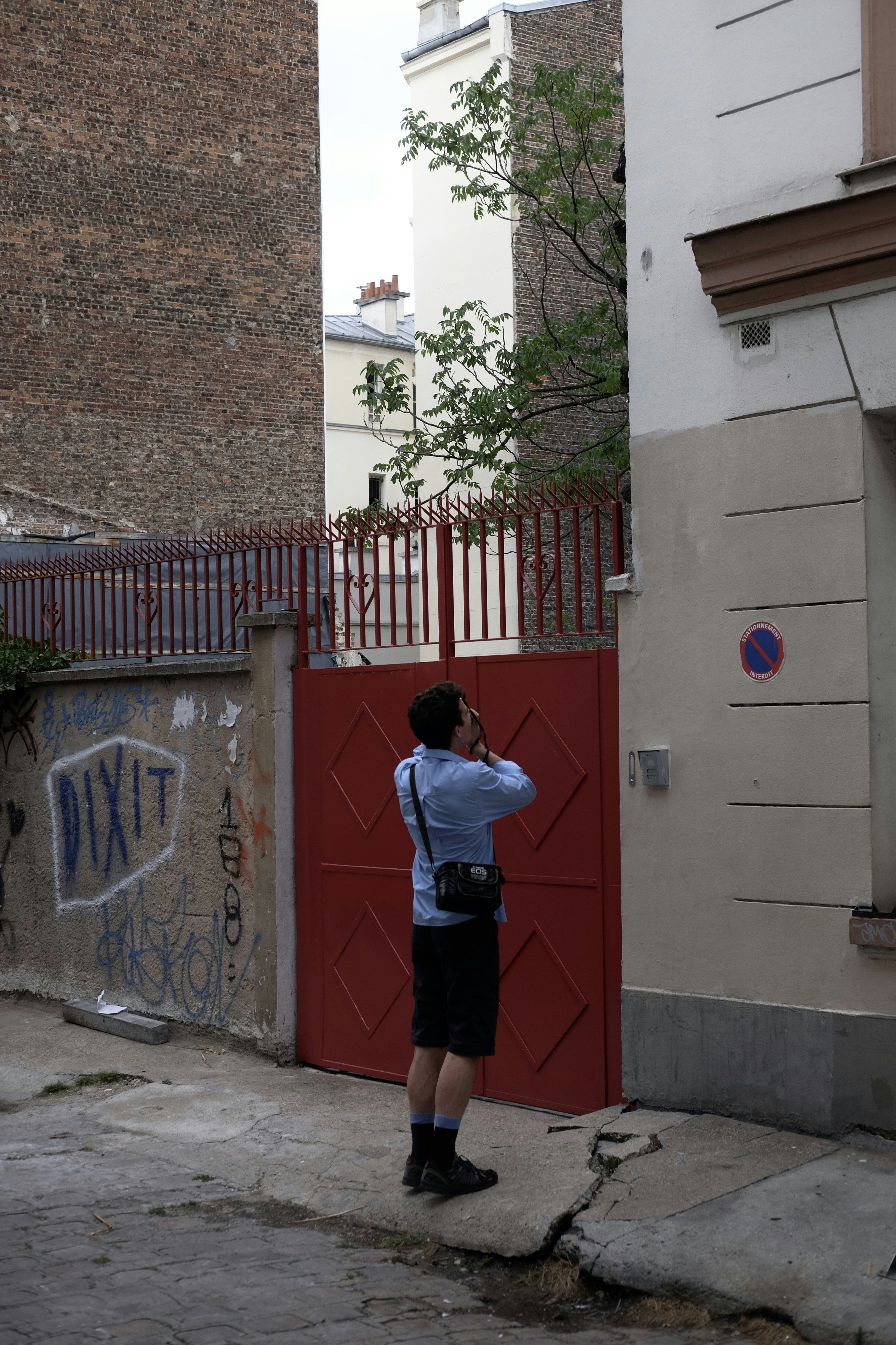 A man in a blue shirt captures a photograph in an alleyway, framed by a red gate and urban graffiti. The scene reflects a blend of architecture and street art.