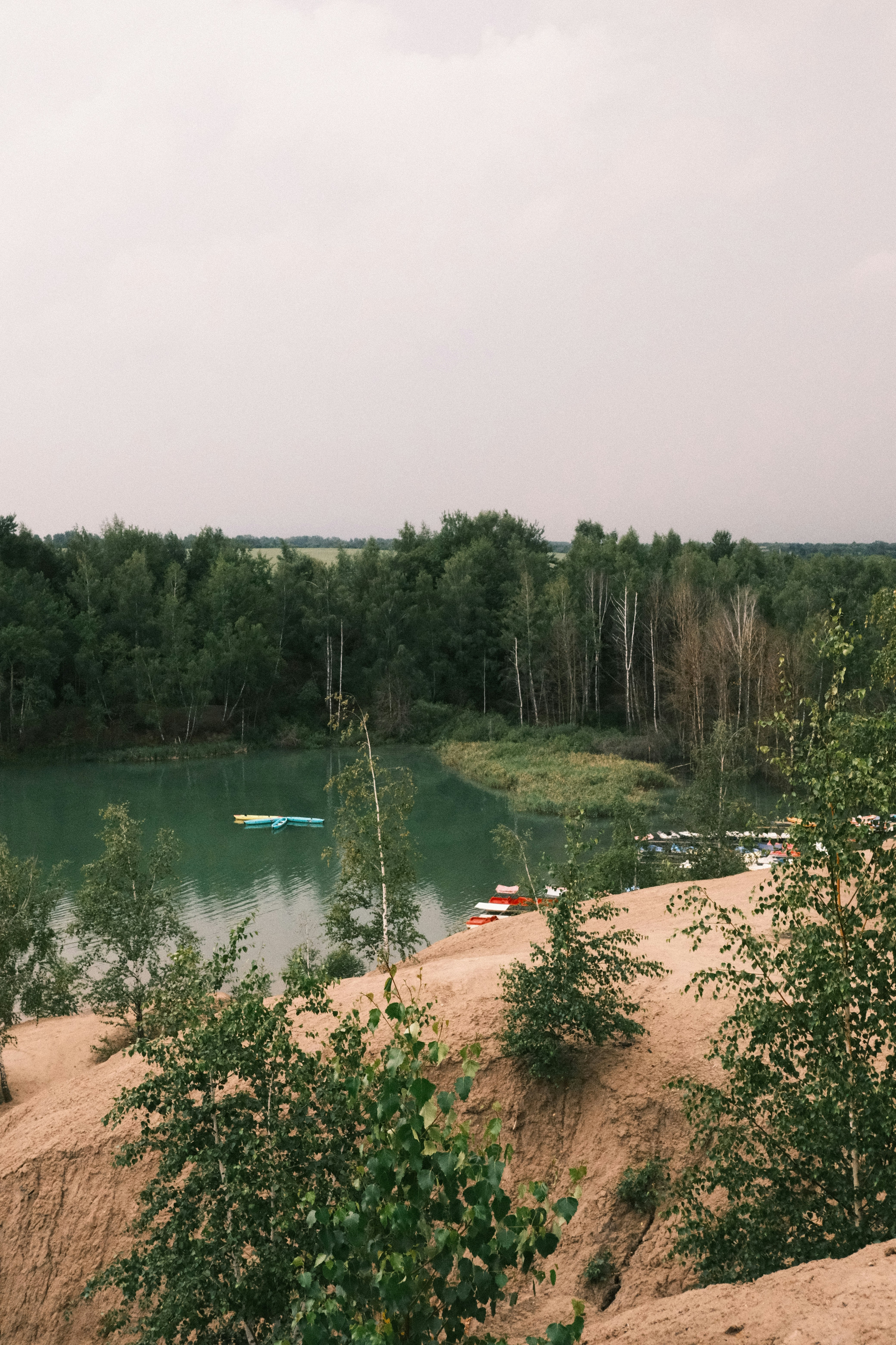 A tranquil river scene featuring colorful kayaks resting along a verdant bank, surrounded by lush trees and a soft, cloudy sky.