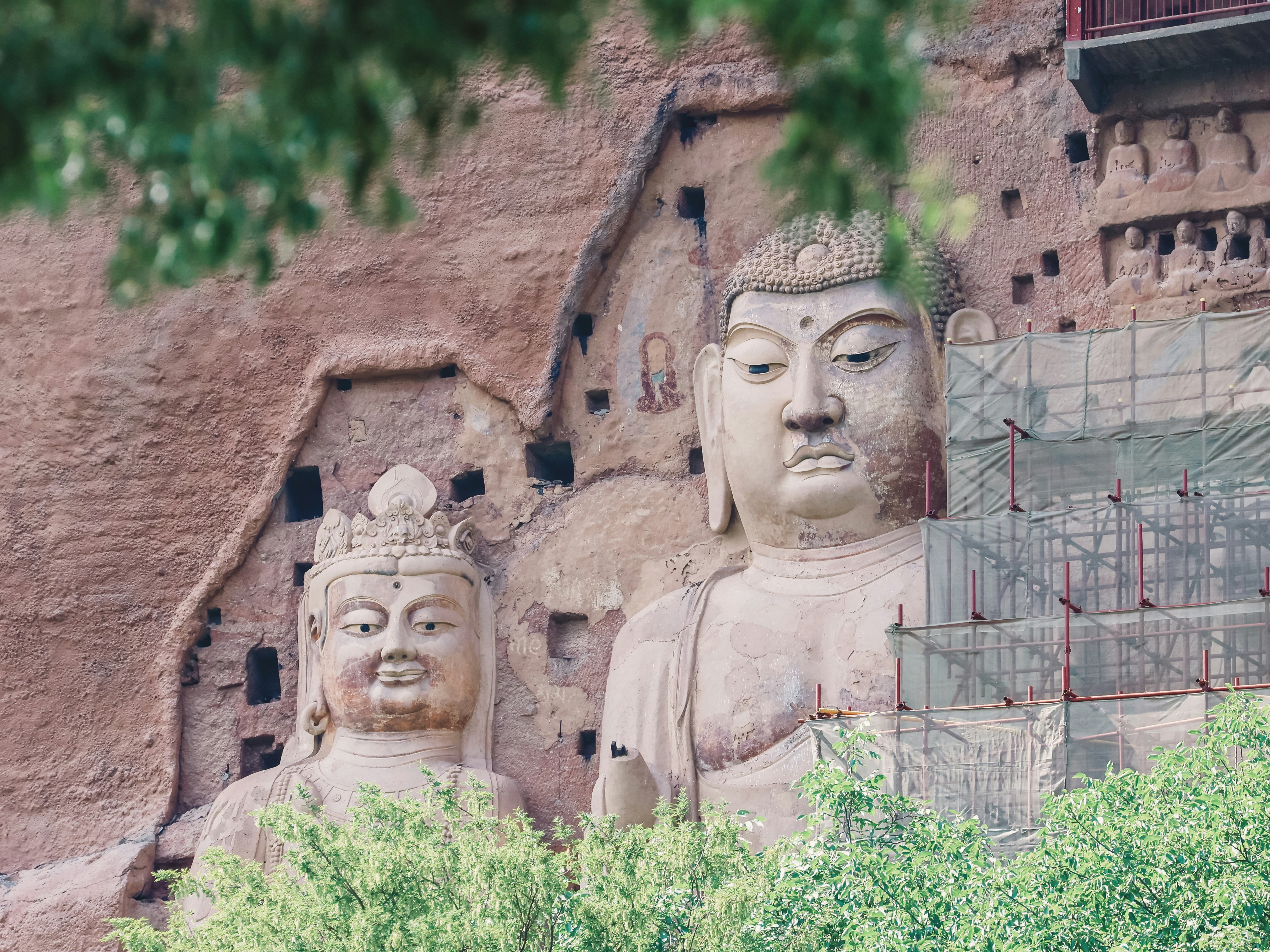 Two majestic Buddha sculptures carved into a cliffside, partially obscured by greenery and scaffolding for restoration work.