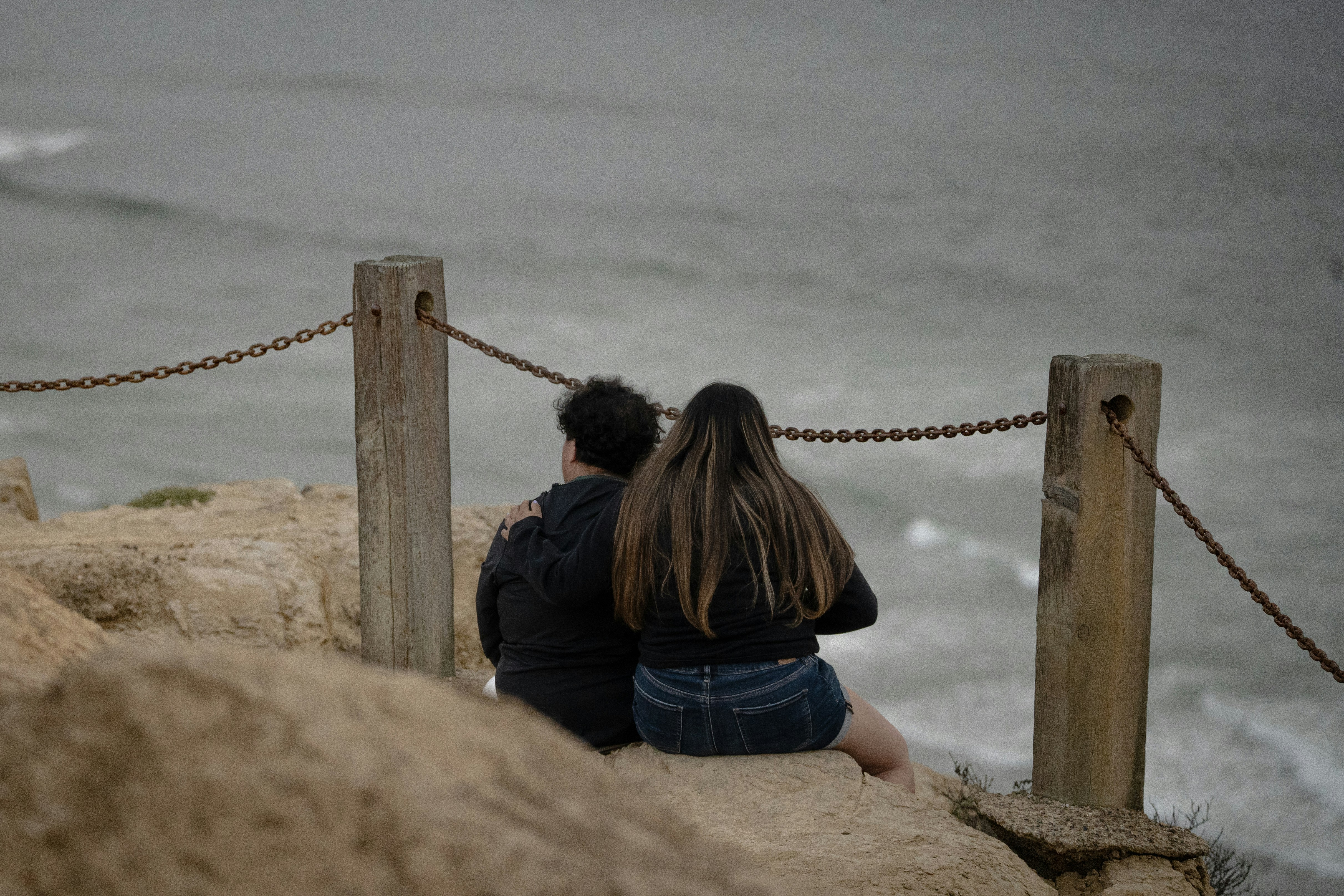 Two individuals sit on a rocky outcrop, gazing at the ocean waves, framed by rustic wooden posts and chains.