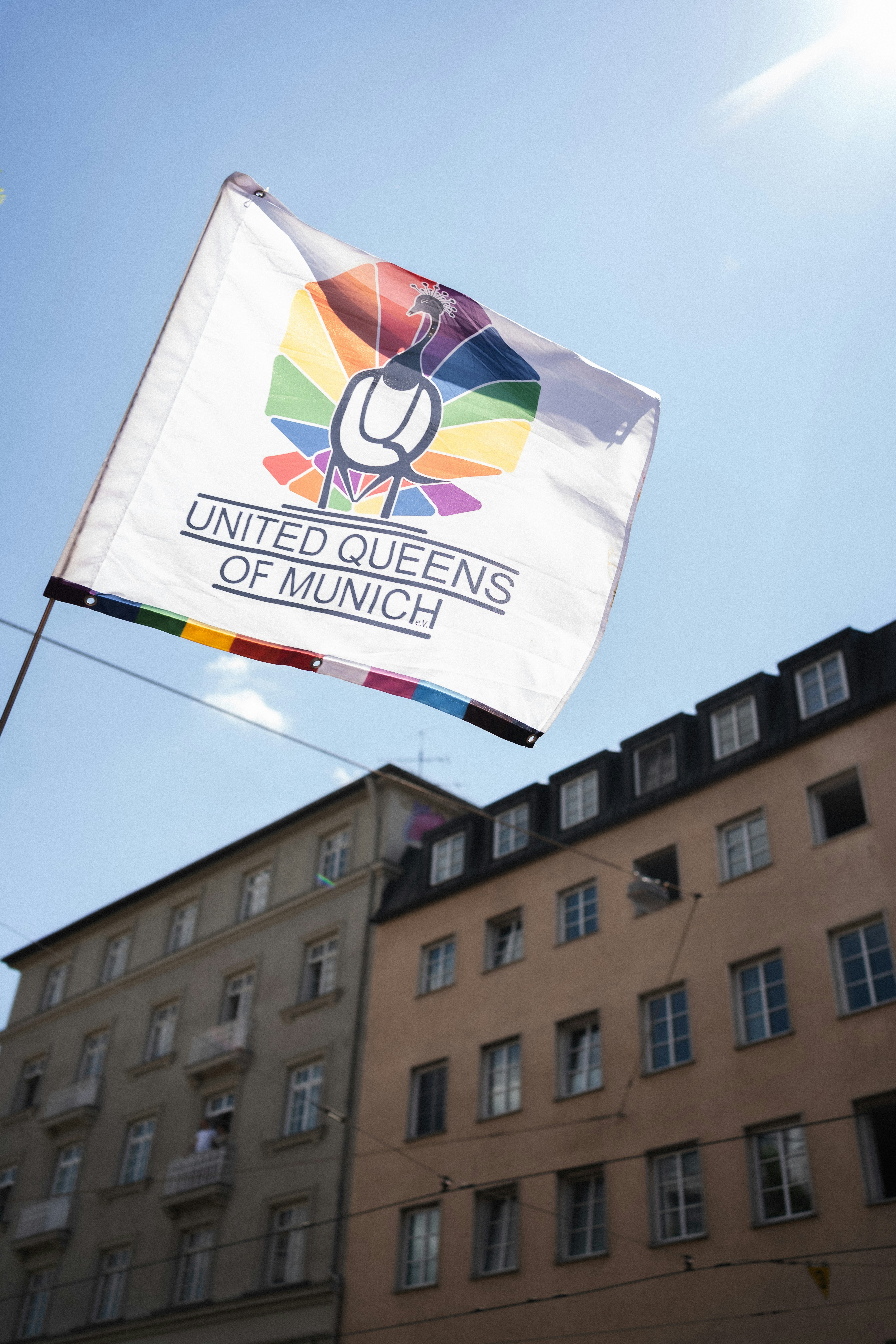 Colorful flag representing the United Queens of Munich, fluttering against a clear blue sky with historic buildings in the background.
