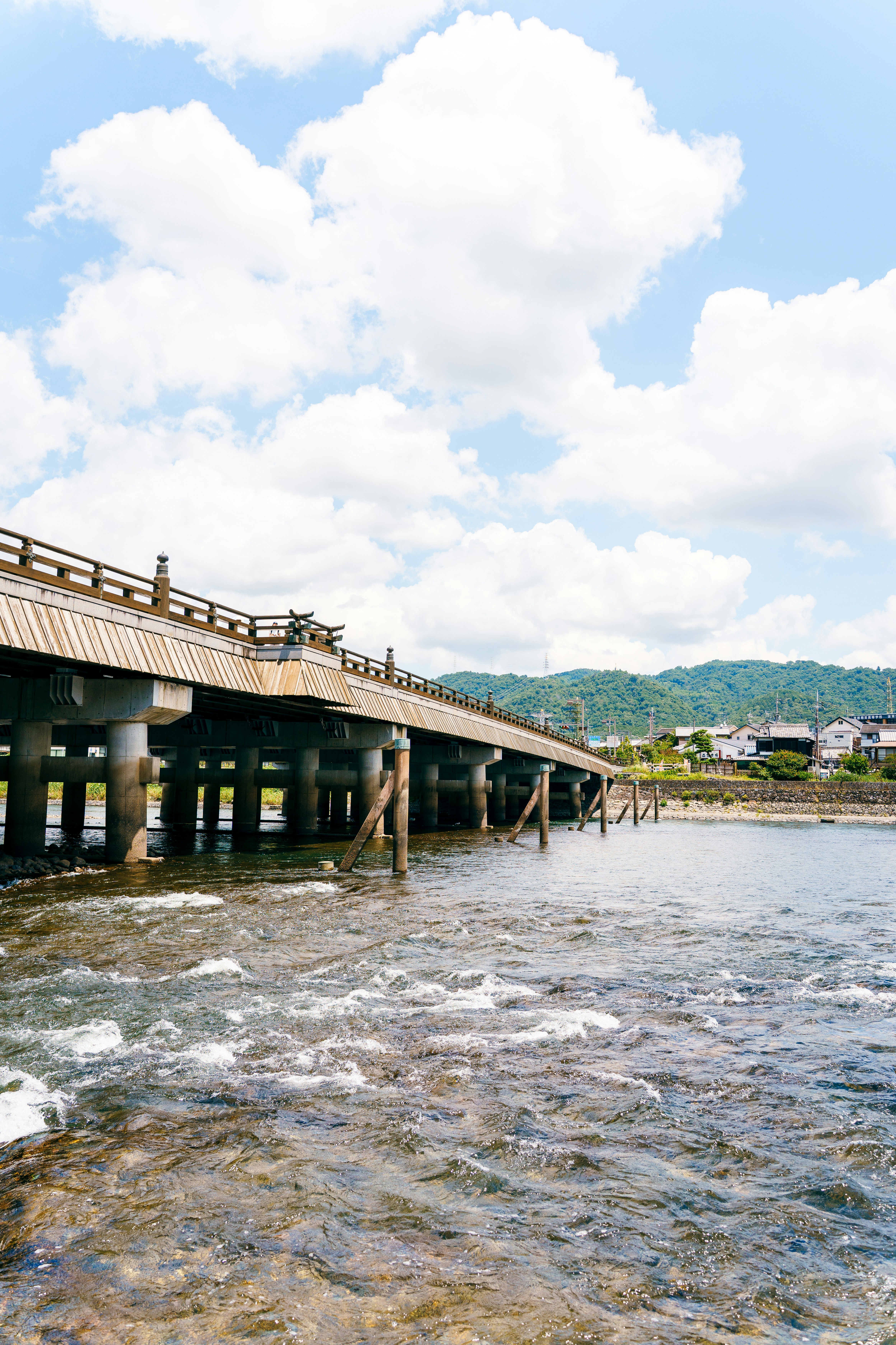 Wooden bridge extending over a flowing river under a bright blue sky dotted with fluffy clouds.