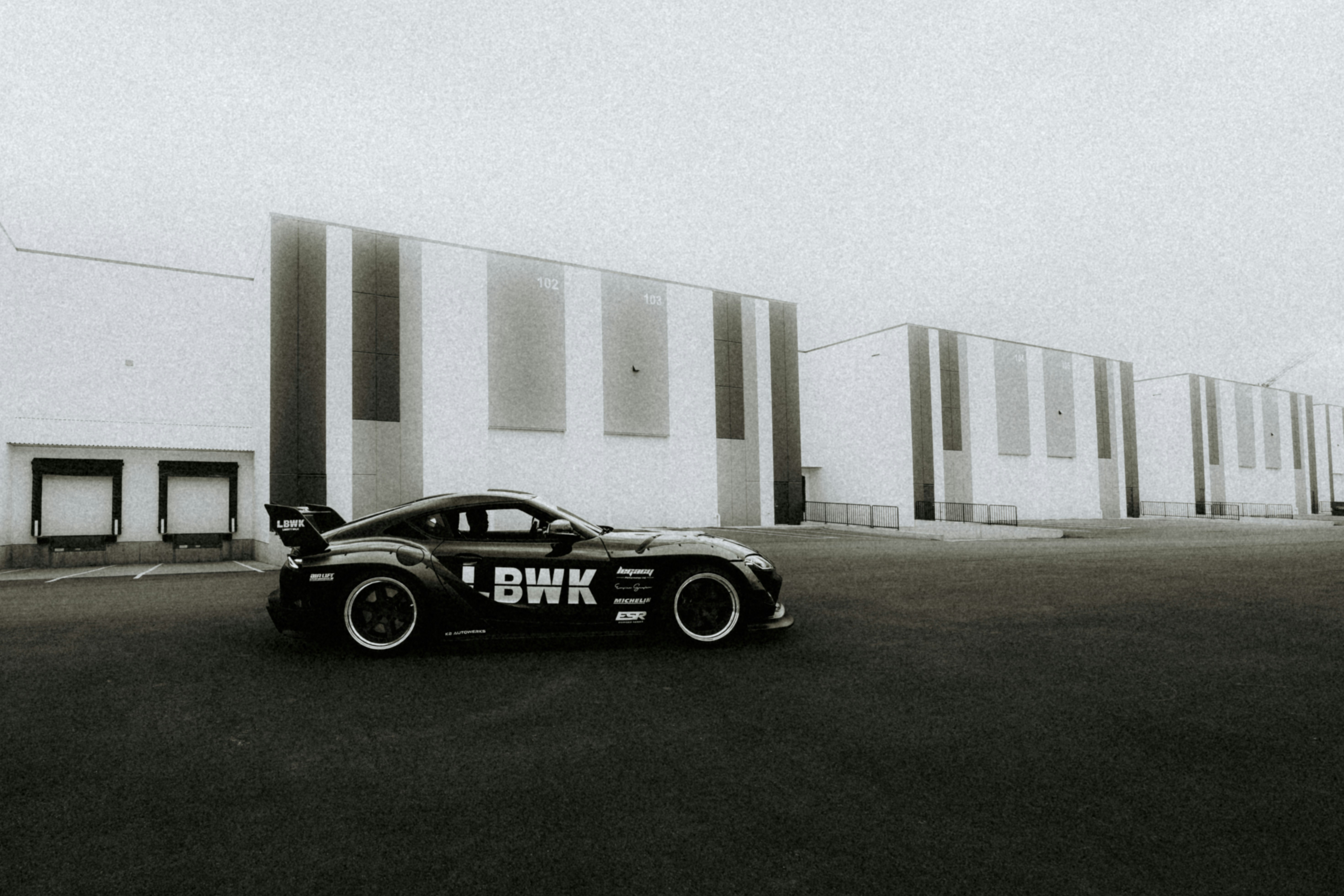 Modified sports car parked in an industrial area, showcasing its sleek design against a backdrop of stark, modern architecture.
