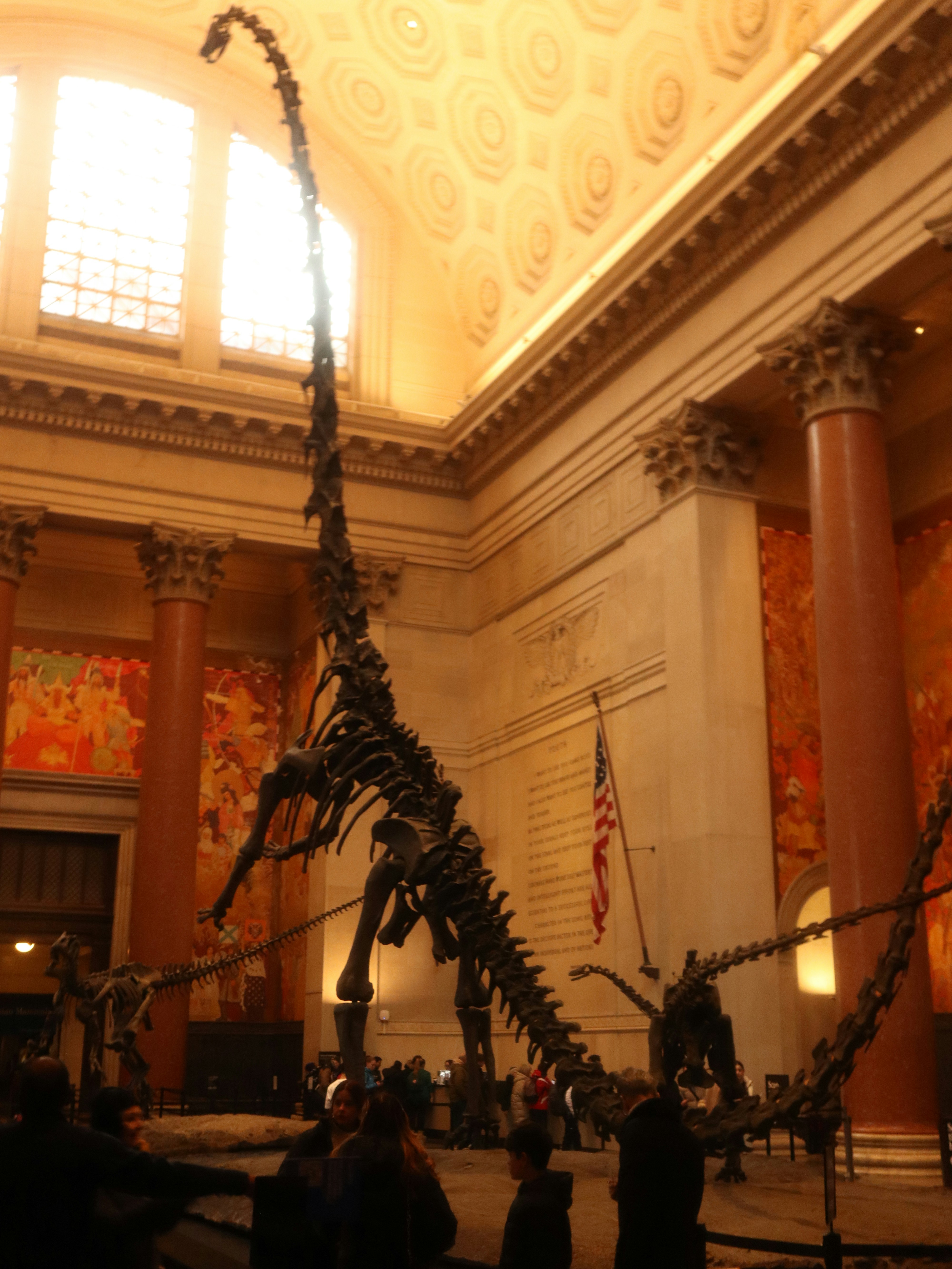 A towering dinosaur skeleton dominates the museum hall, surrounded by visitors admiring its grandeur. The ornate architecture and vibrant murals enhance the historical ambiance.